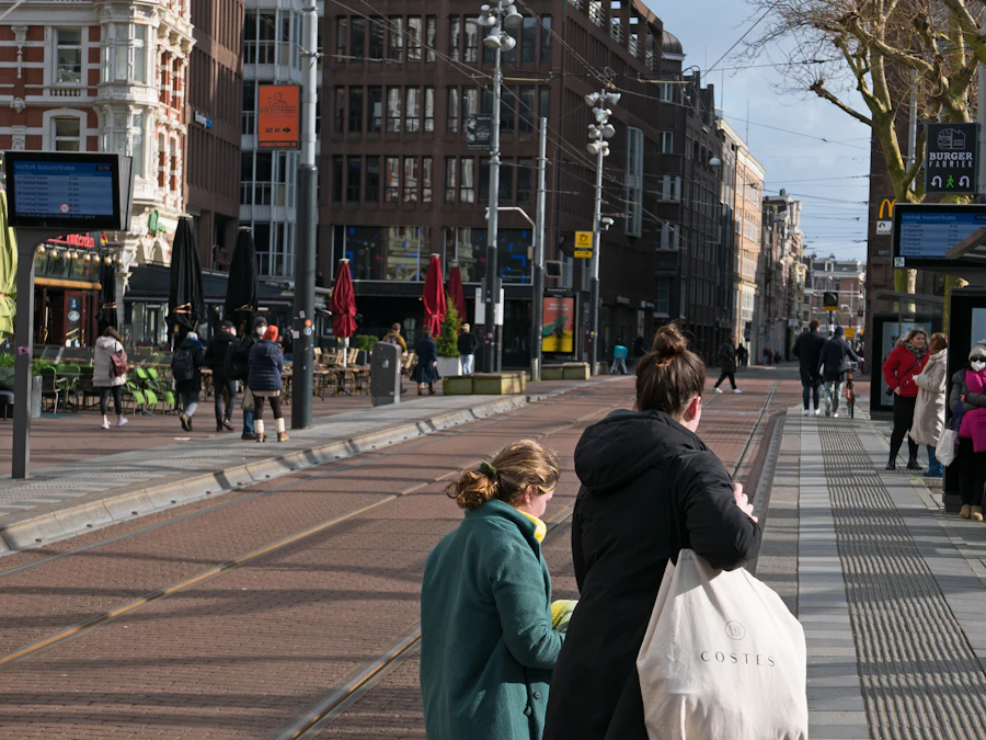 Two people walk along tram tracks on a busy city street lined with shops, cafes, and signs. Pedestrians and buildings recede into the distance under bright daylight.