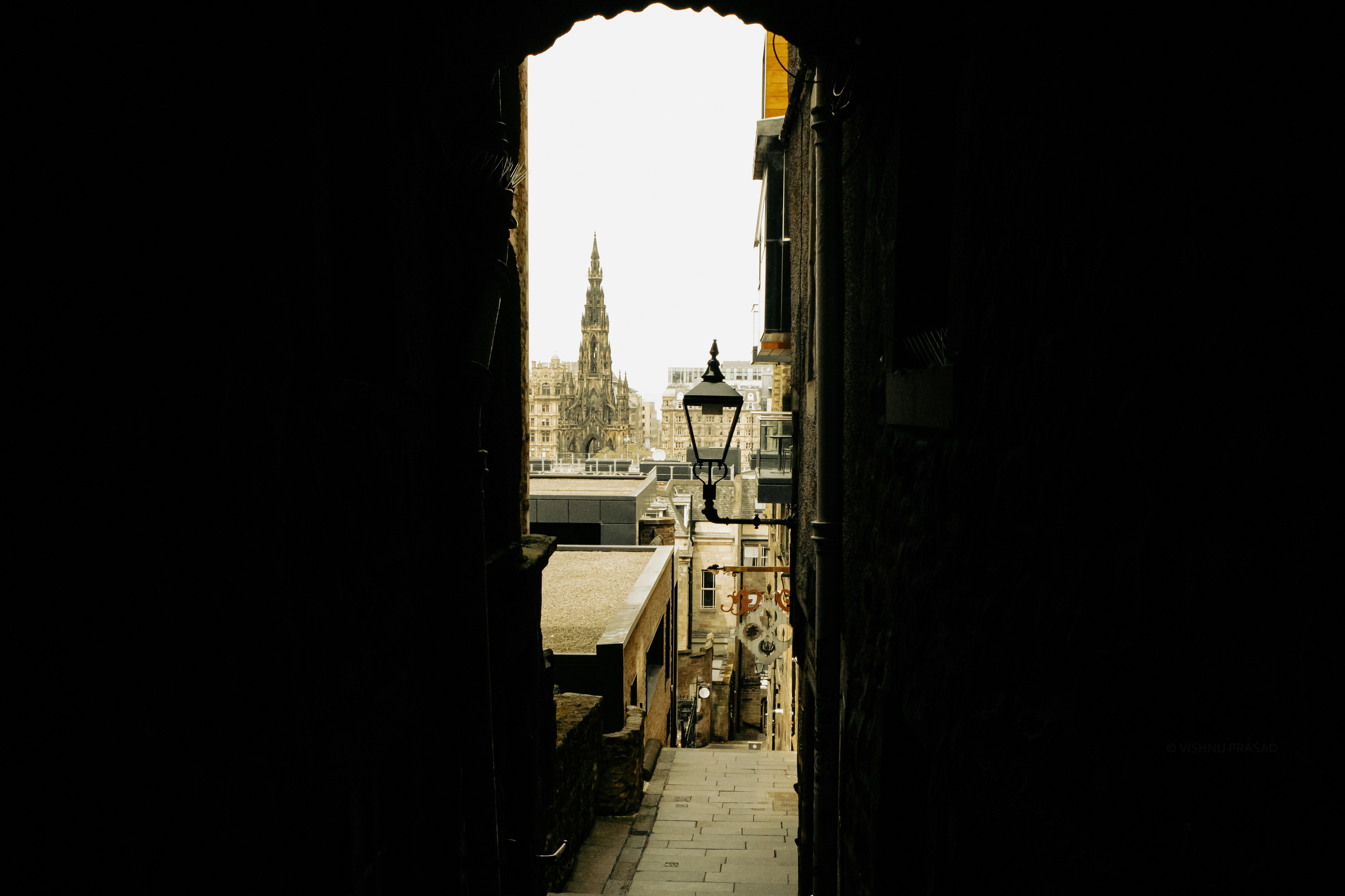 Narrow alleyway opens to a view of an old town with a distant church spire under a cloudy sky.