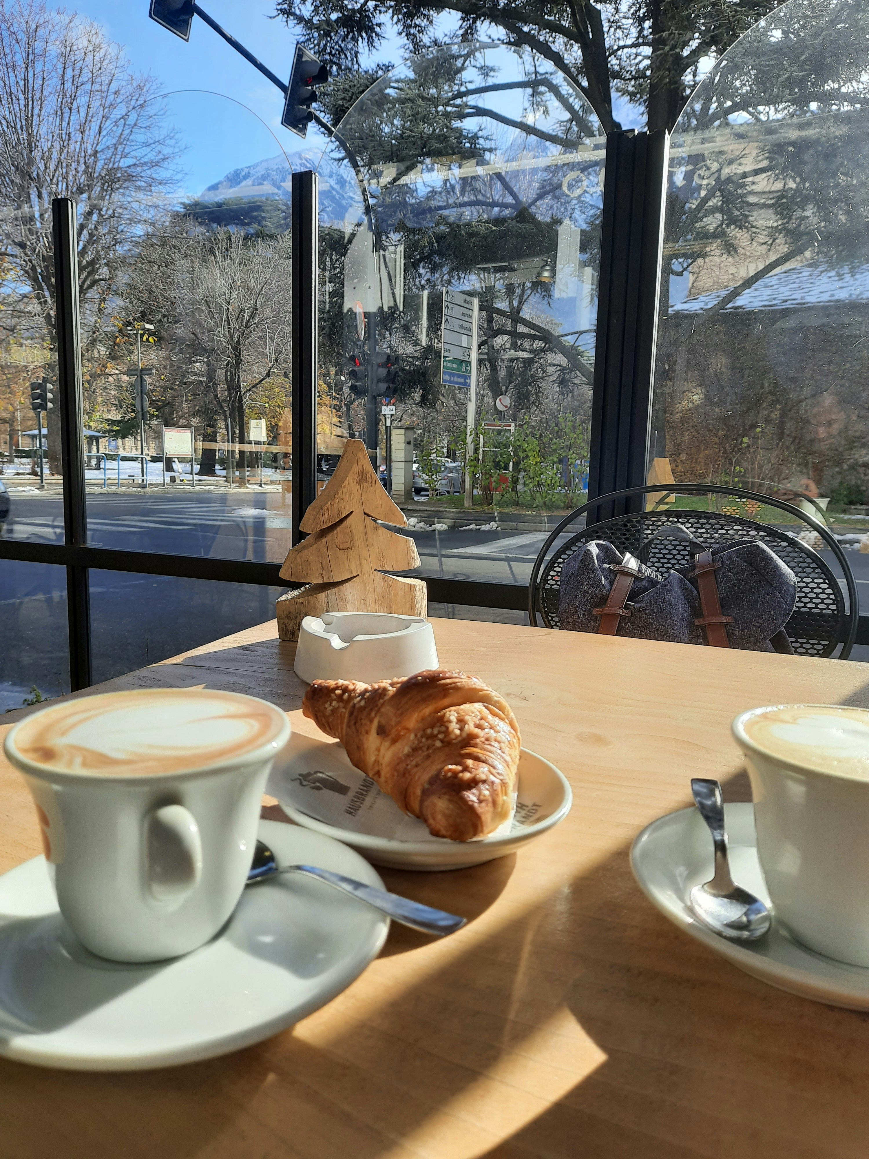 Photograph of a sunlit café table with a croissant on a plate and two cups of coffee, viewed through a glass window. A wooden decorative tree sits at the center.