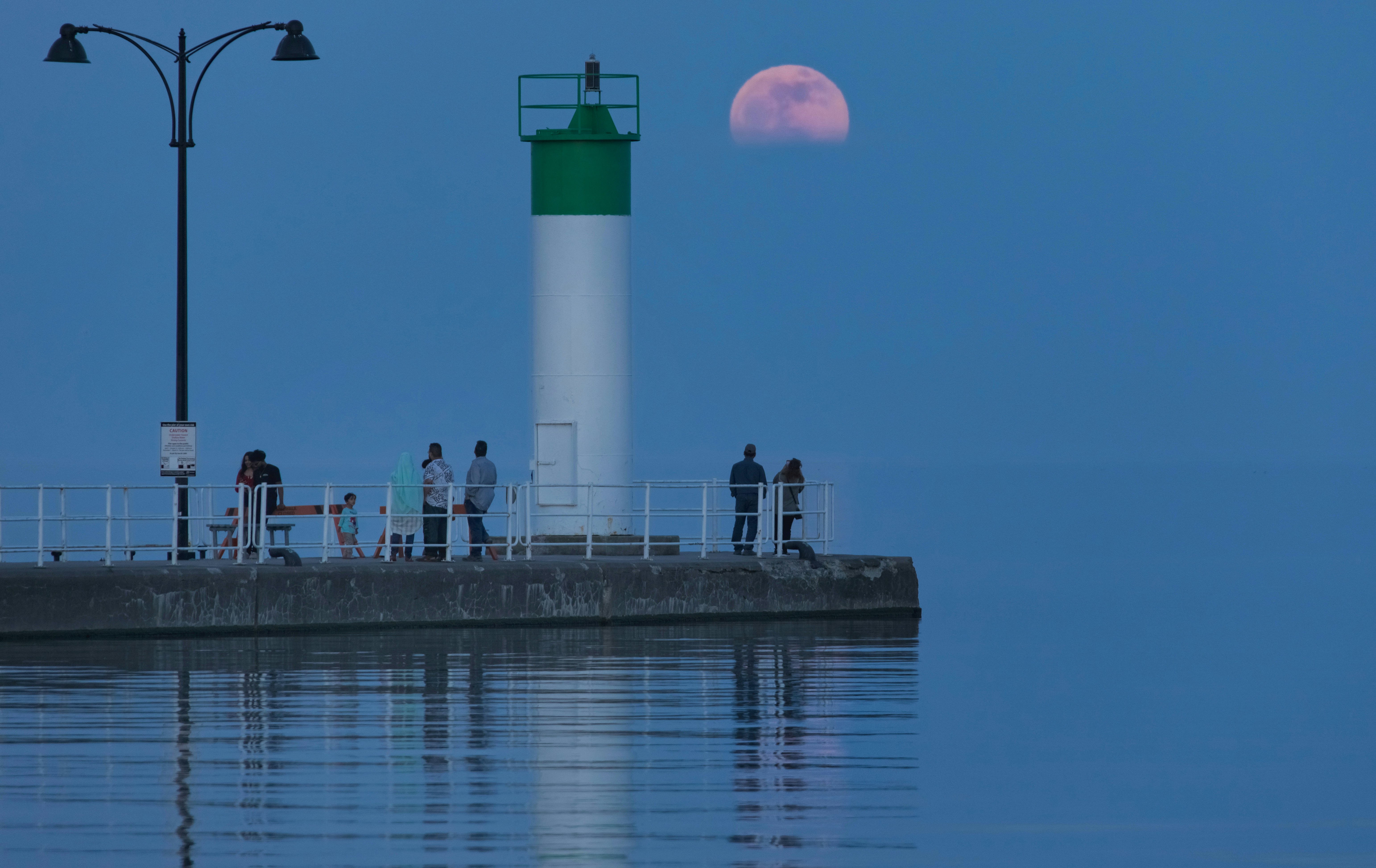 Moon rising behind a lakeside pier with a green and white lighthouse and silhouetted figures against a twilight sky.
