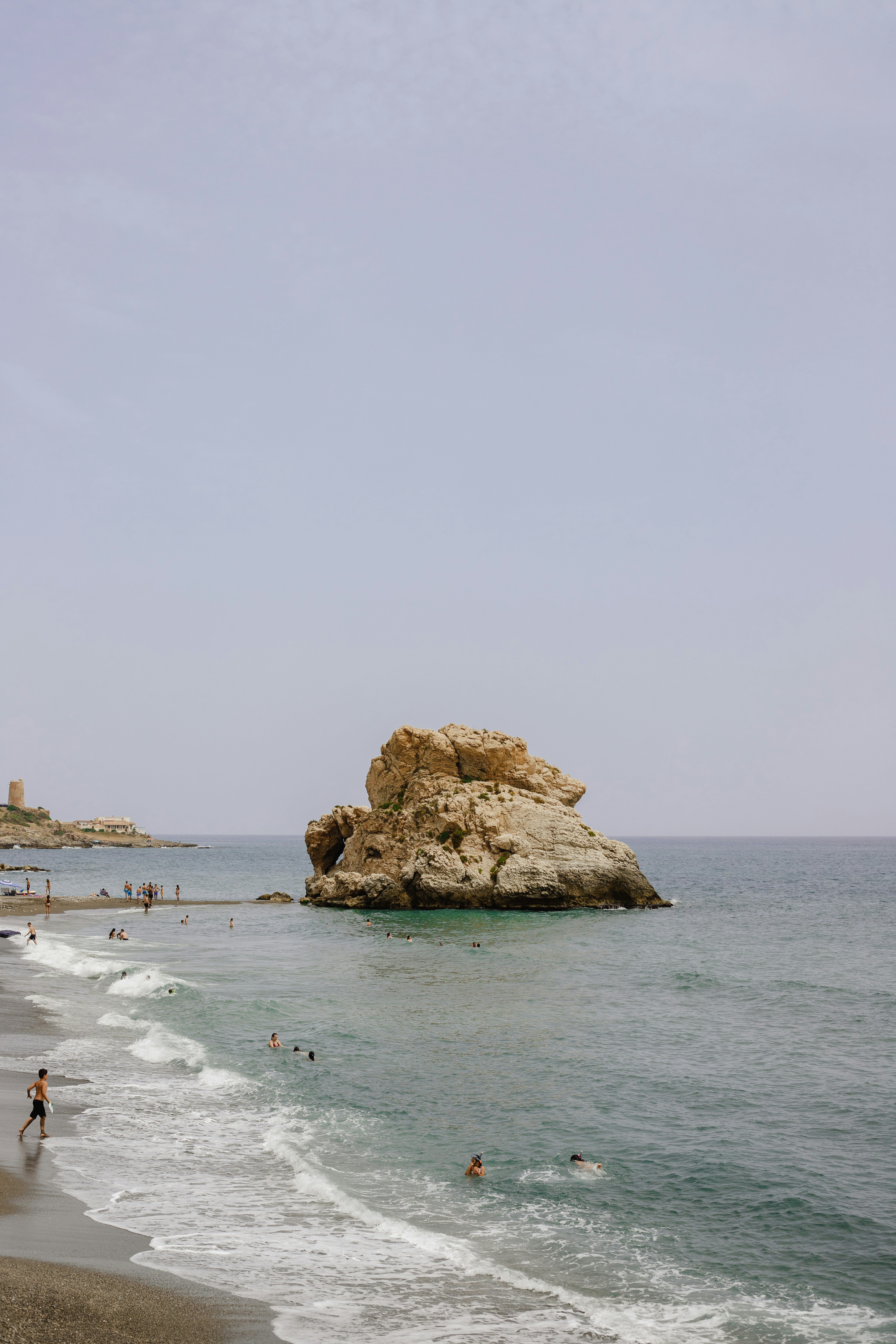 A rocky outcrop stands majestically in the gentle waves, surrounded by beachgoers enjoying the sun and surf. The tranquil sea reflects the soft hues of the sky.