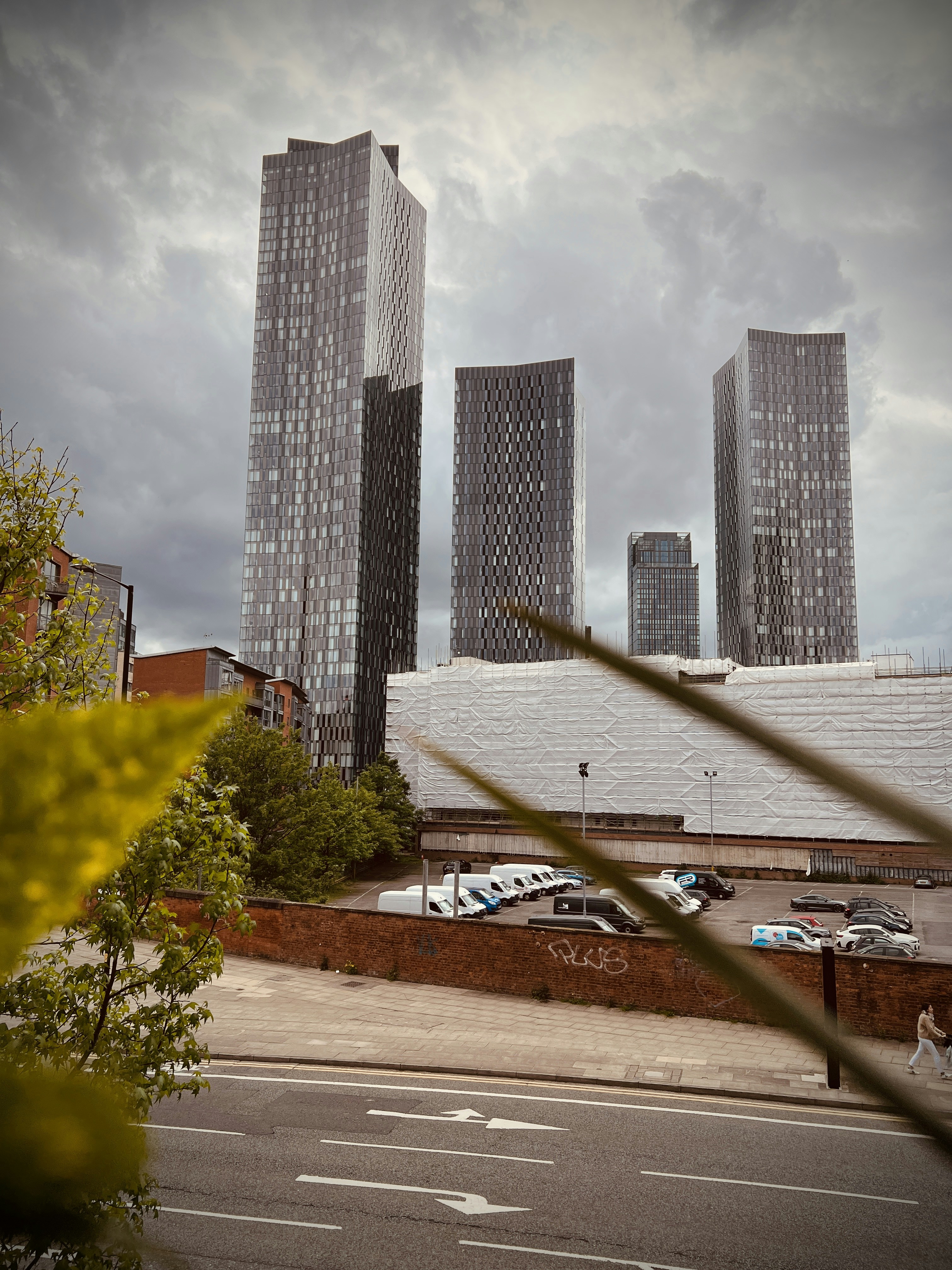 Four sleek skyscrapers rise against a moody sky, framed by greenery in the foreground. The juxtaposition of nature and modern architecture highlights urban development.