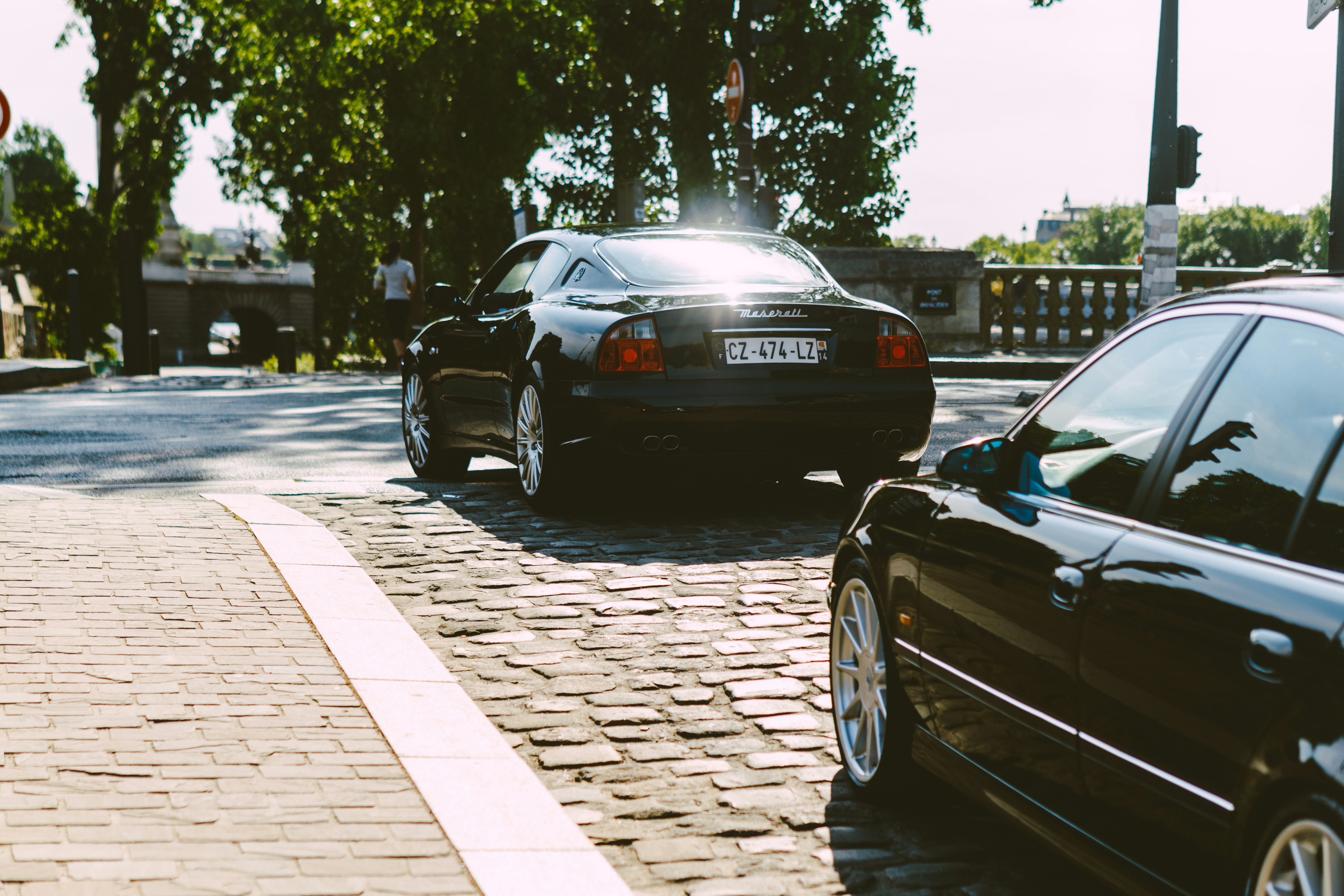 Two sleek black cars navigating a sunlit cobblestone street, framed by lush greenery and urban scenery.