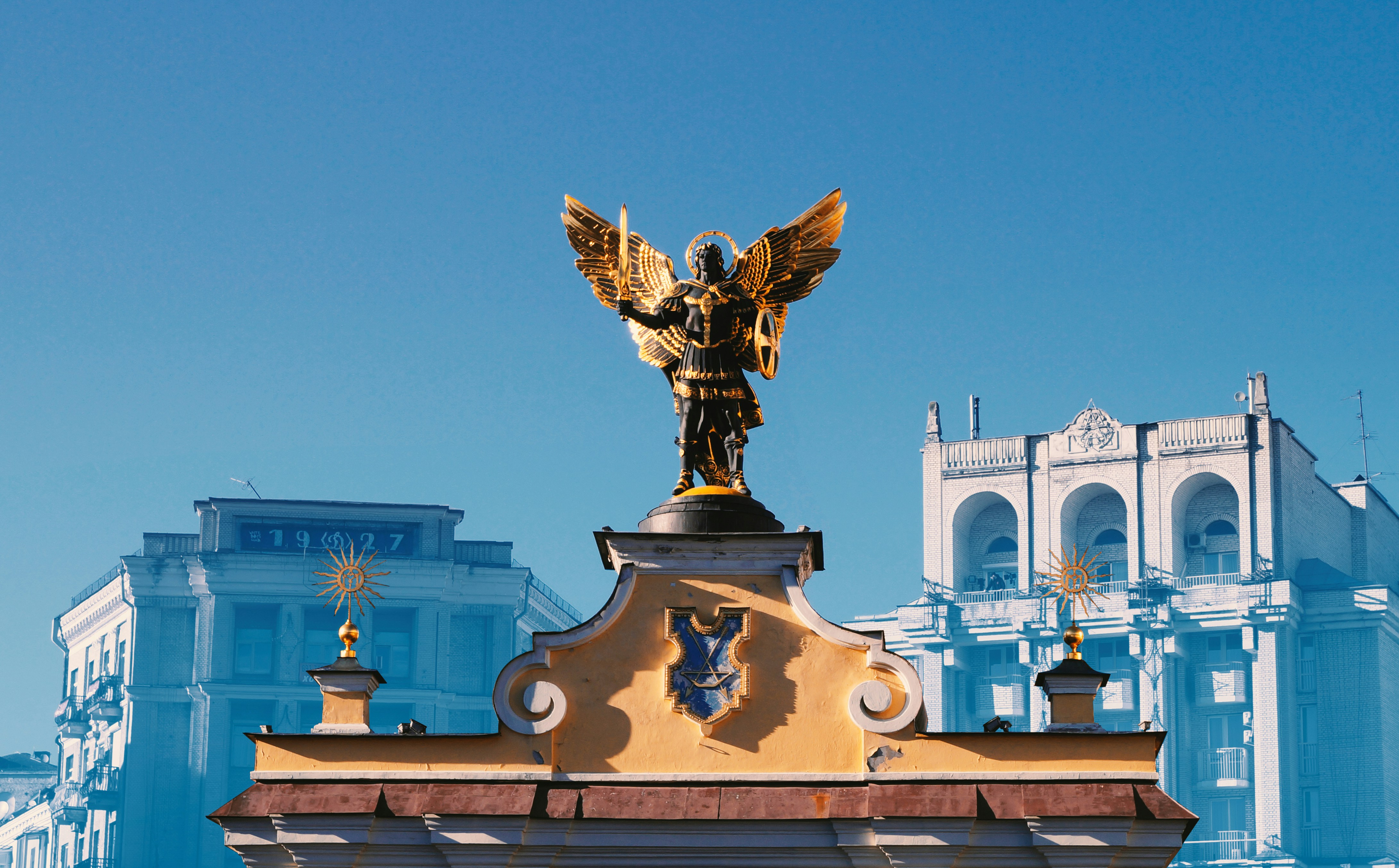 Photograph of a gilded winged statue atop an ornate pediment, with a pale orange façade and a clear blue sky.