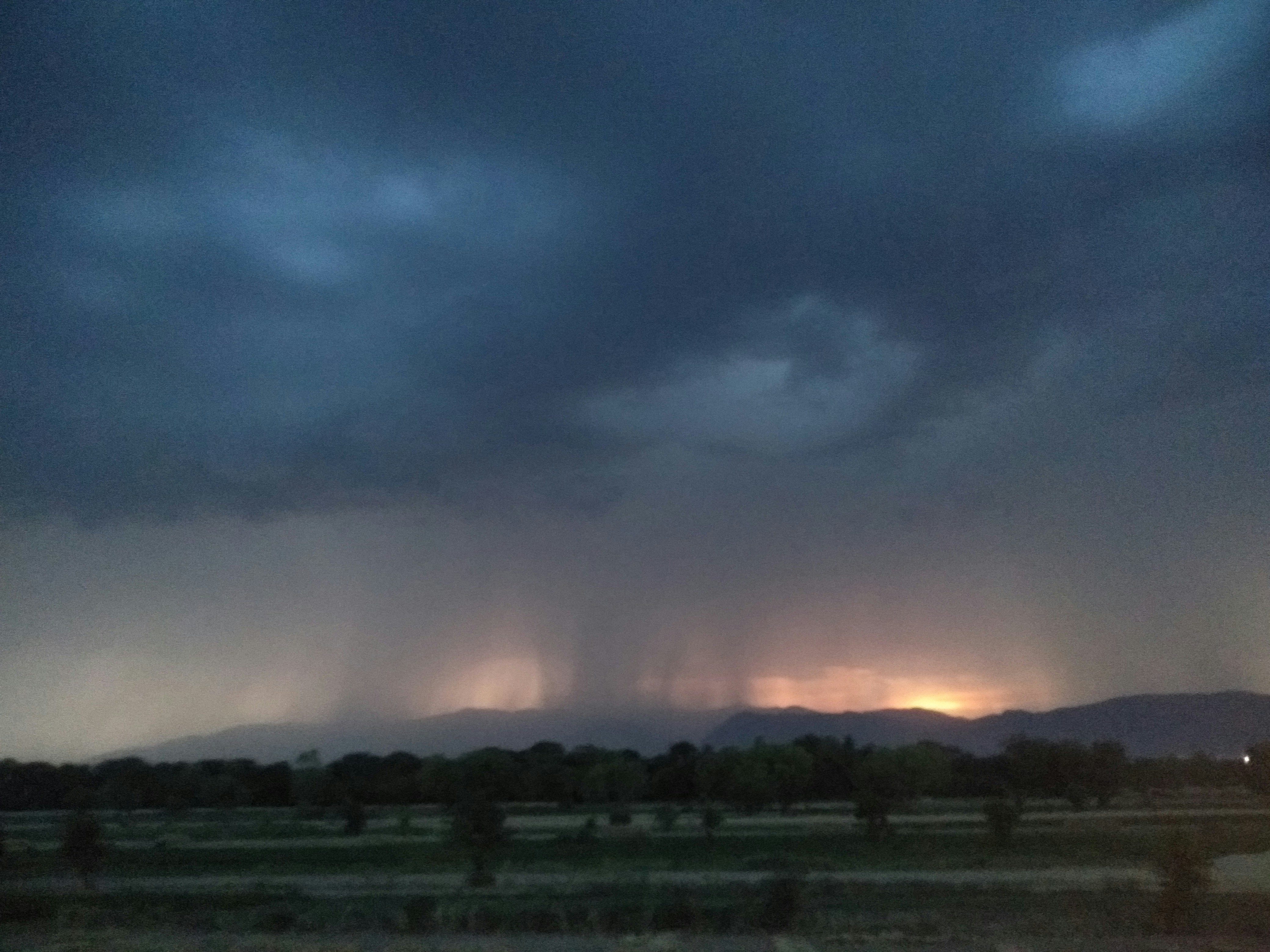 Dramatic clouds loom over distant mountains, with rain cascading down and a faint glow on the horizon hinting at the setting sun.