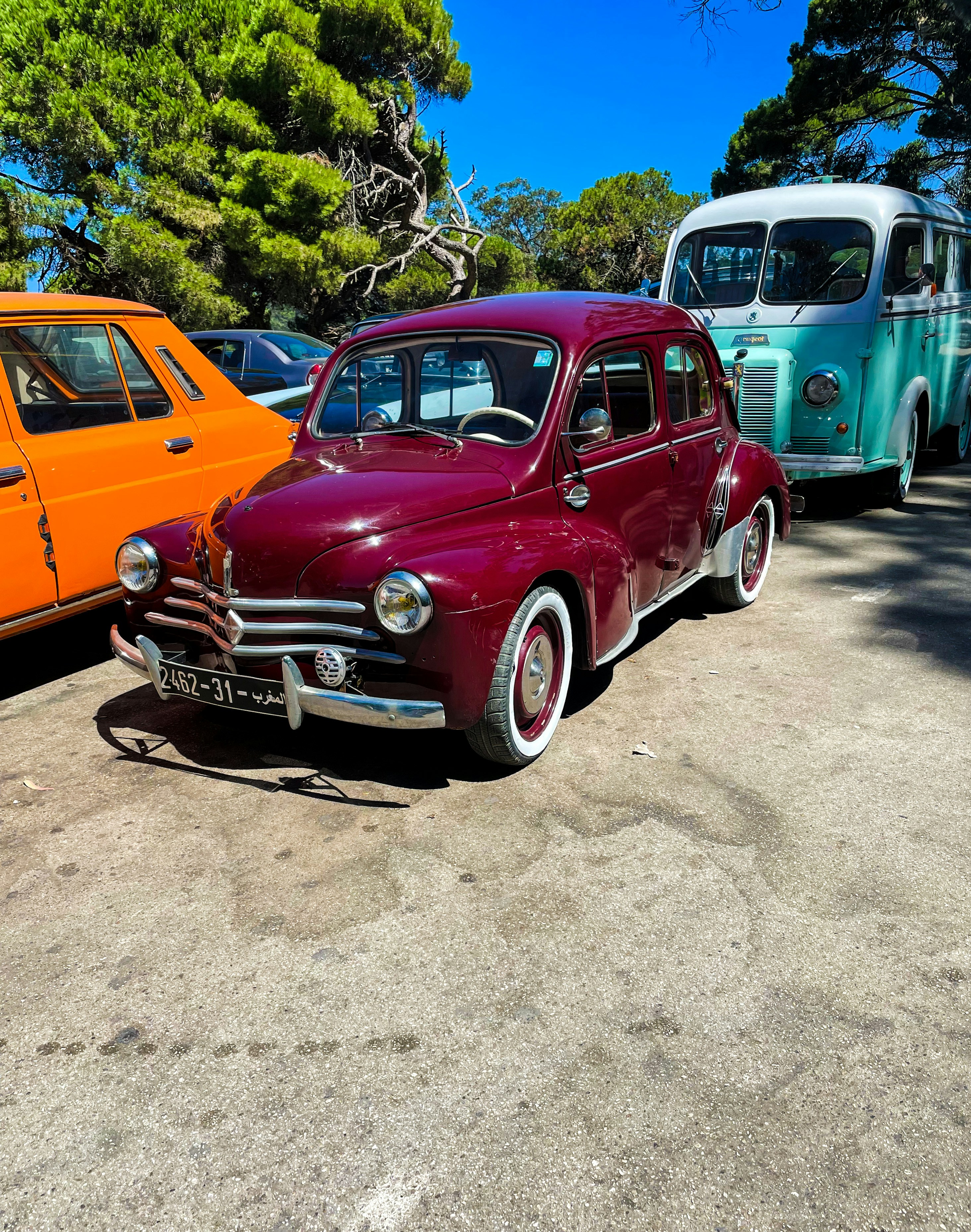 A maroon vintage sedan sits front and center in a sunlit car lot, flanked by an orange classic on the left and a teal bus in the background.