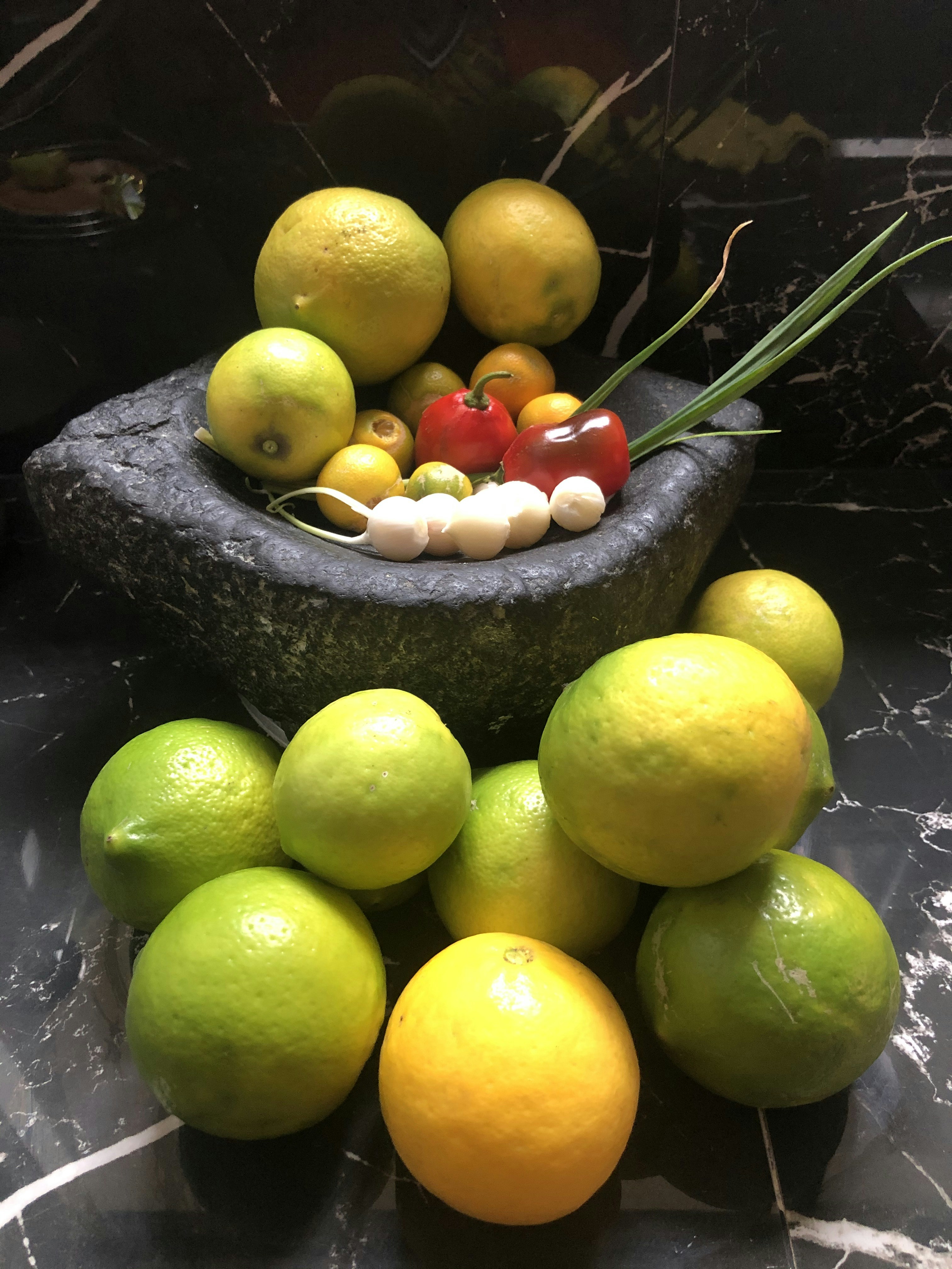 A beautifully arranged collection of various citrus fruits and vegetables in a textured stone bowl, showcasing vibrant colors against a dark marble background.