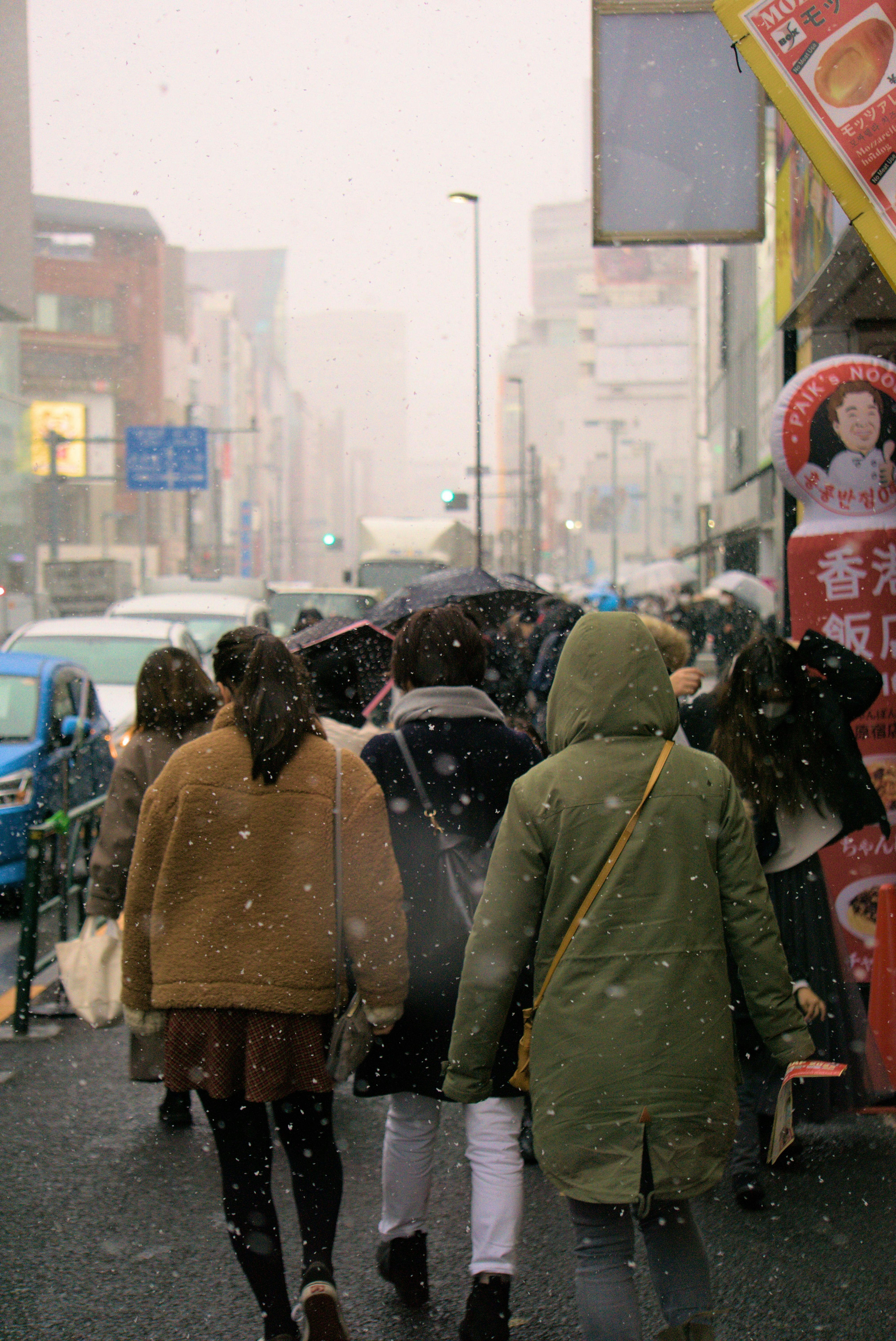 A group of people walking in a snowy urban environment, with umbrellas shielding them from the falling snow. The scene captures the essence of a bustling city day in winter.