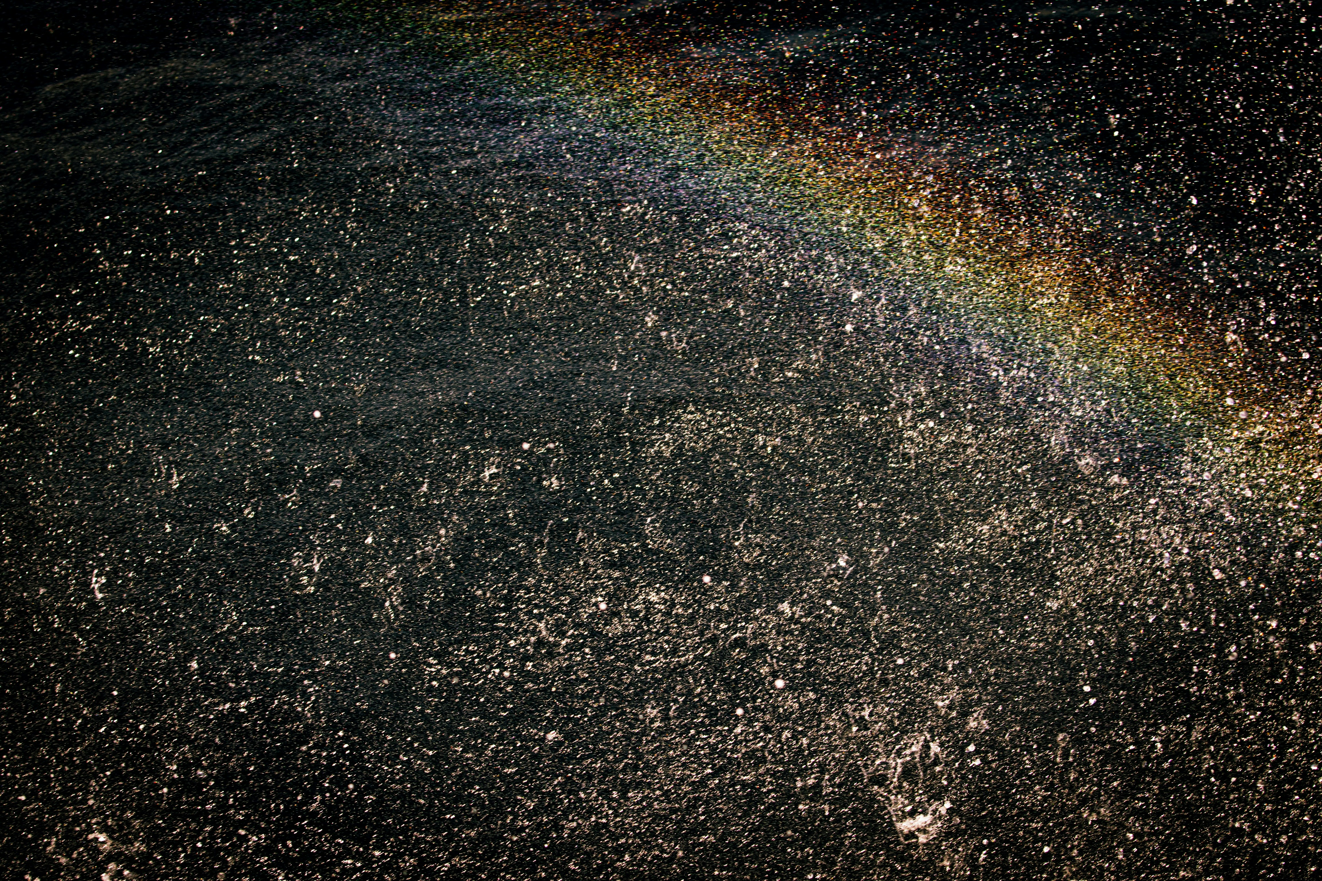 Macro photograph of countless dust specks on a dark surface, with a faint rainbow arc sweeping across the frame.