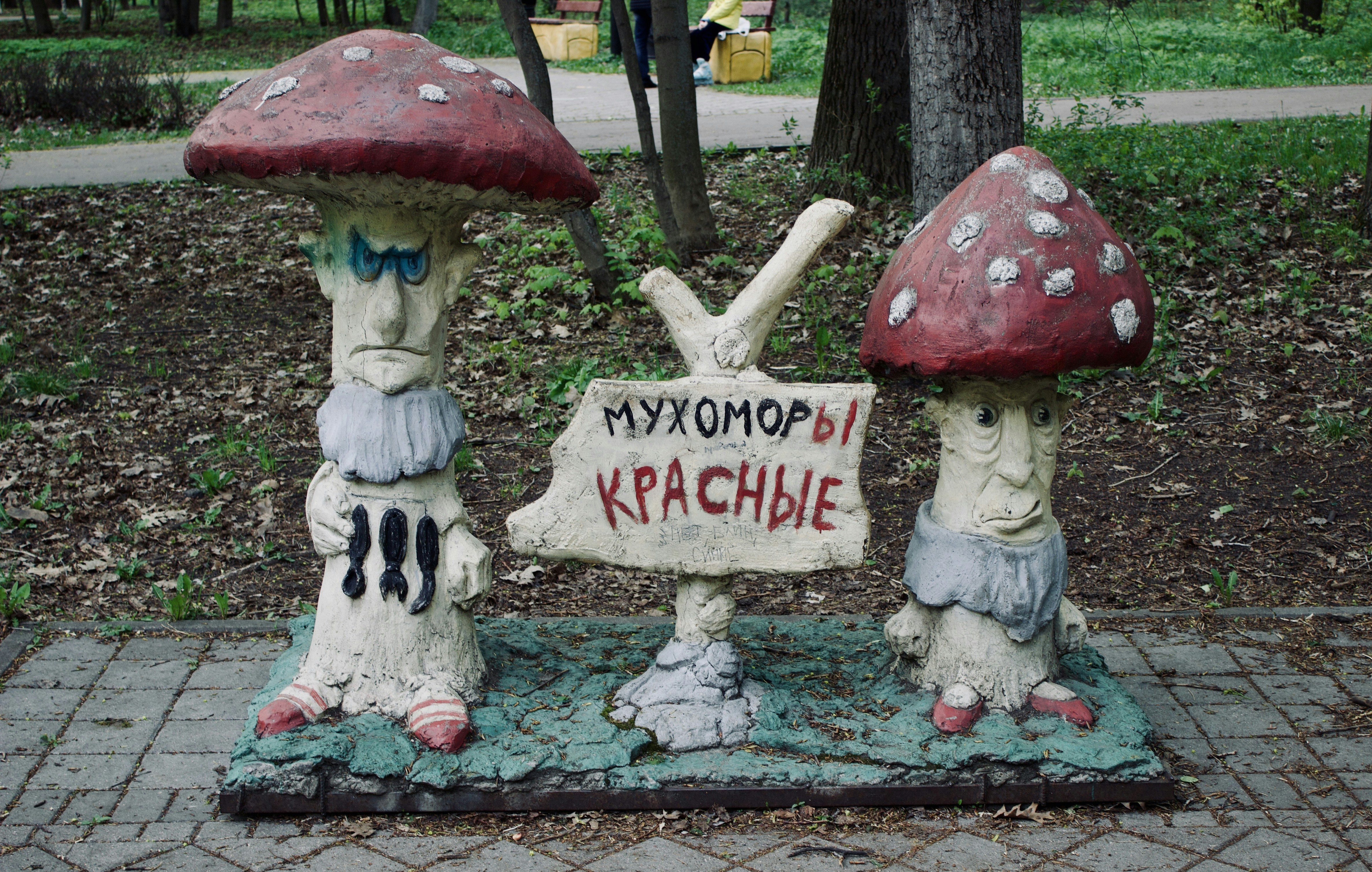 Two cartoonish mushroom sculptures with expressive faces stand beside a sign, inviting visitors to explore the world of red mushrooms. The vibrant colors and playful design add charm to the park setting.