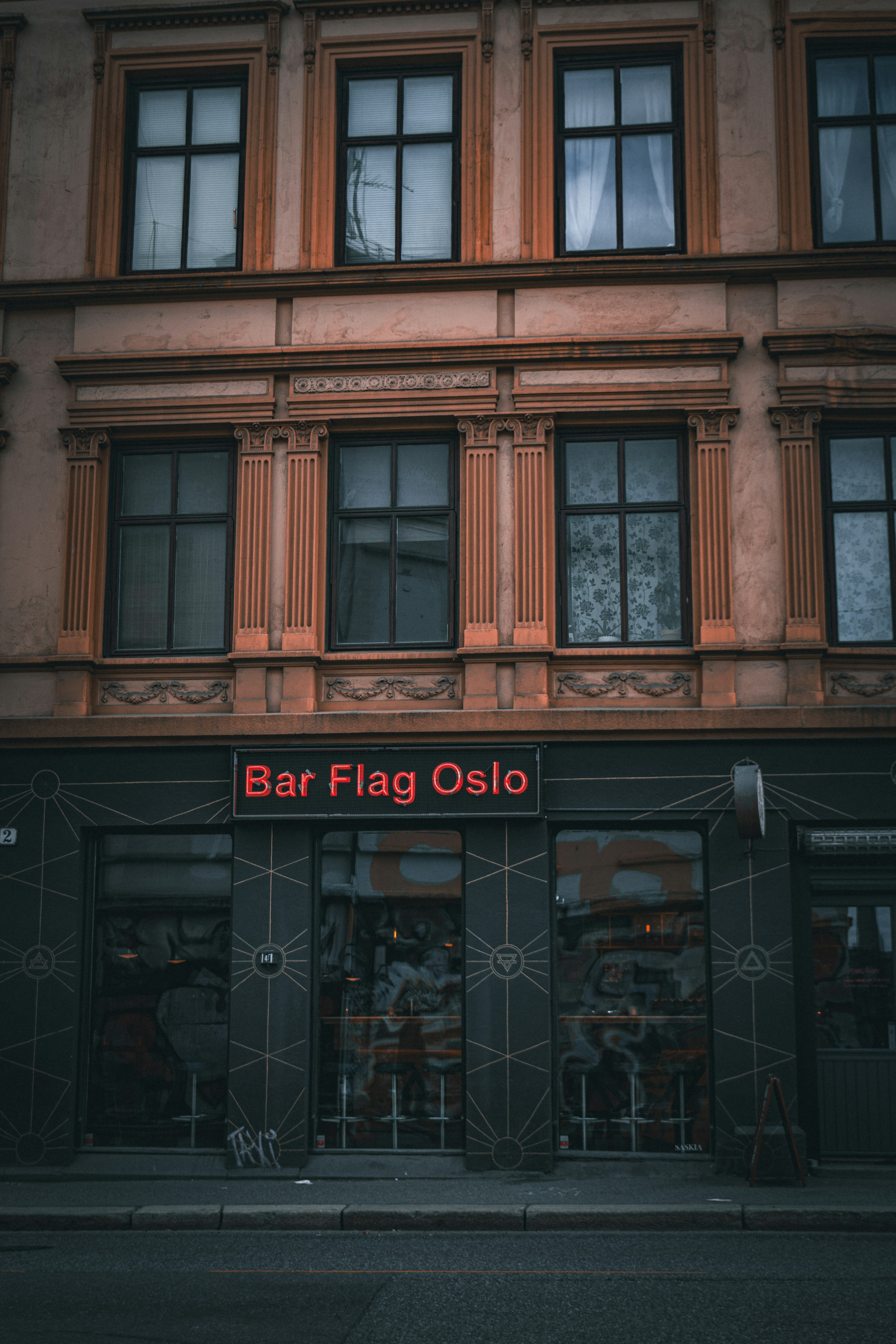 Facade of Bar Flag Oslo showcasing ornate architectural details and illuminated signage. The contrast between the building's classic features and modern bar aesthetics is striking.
