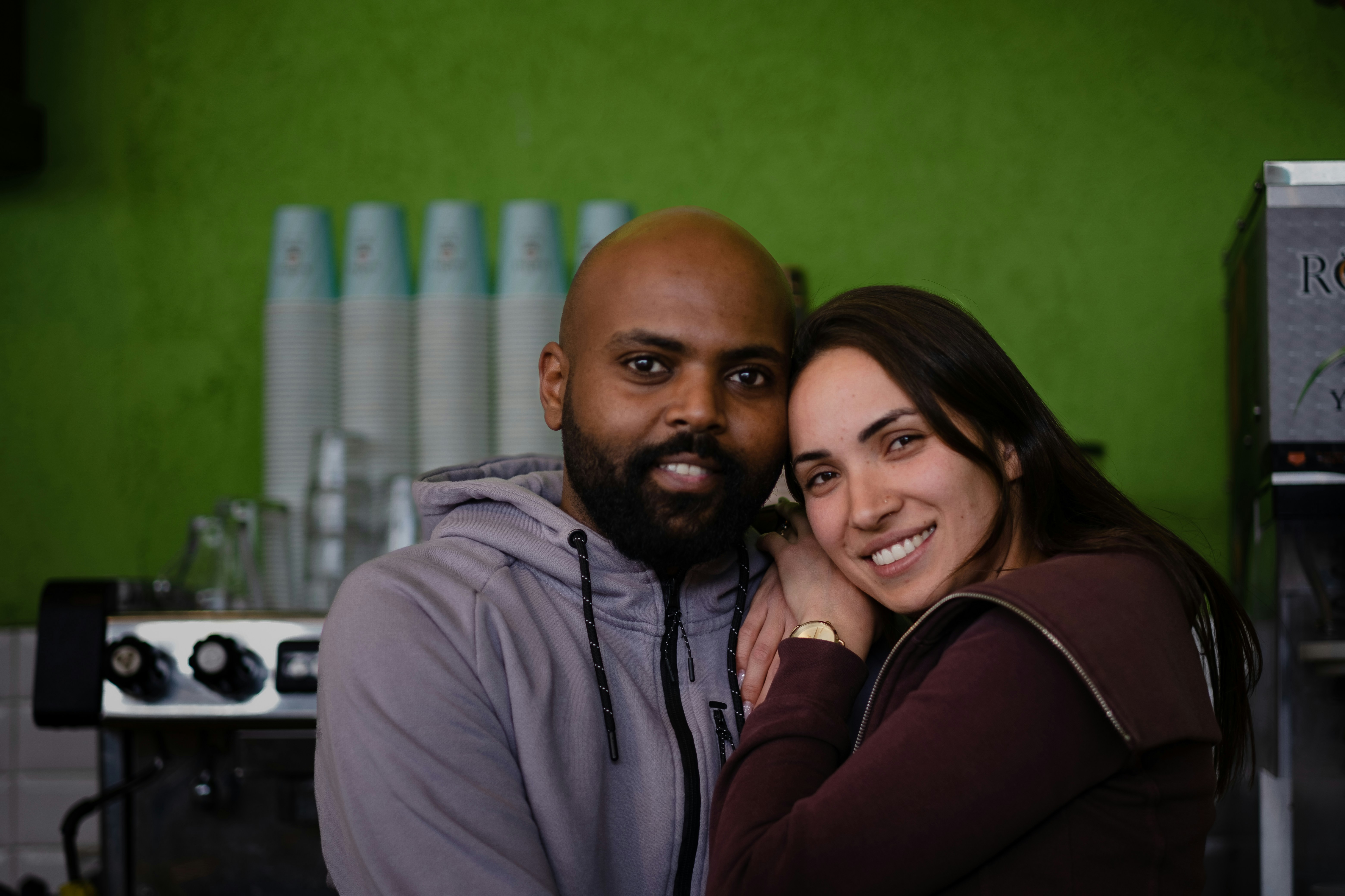 Couple sharing a warm embrace against a vibrant green café wall with coffee machines in the background.