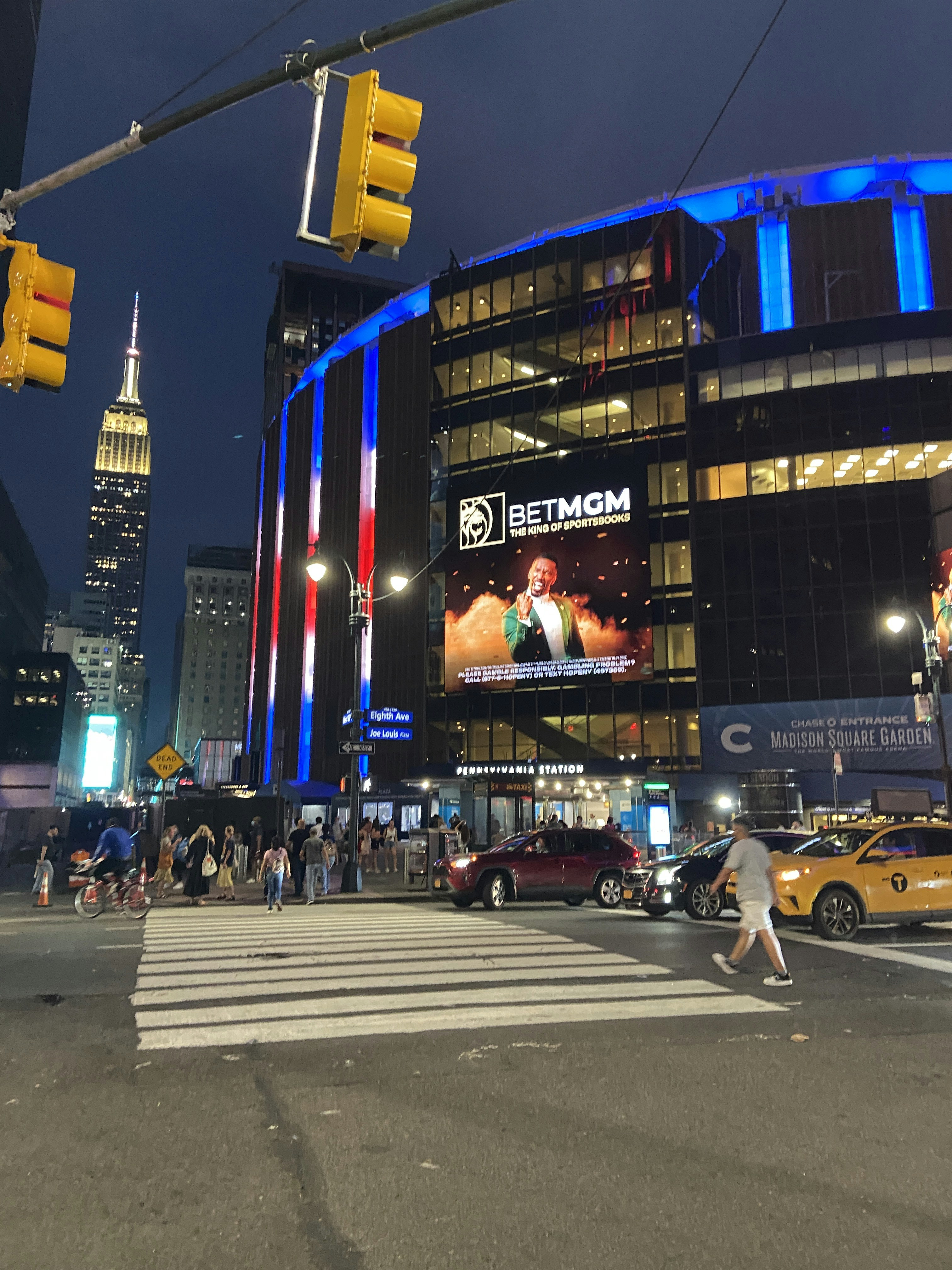 Vibrant city scene showcasing Madison Square Garden illuminated by colorful lights, with the Empire State Building in the background. A bustling crosswalk adds to the urban energy.