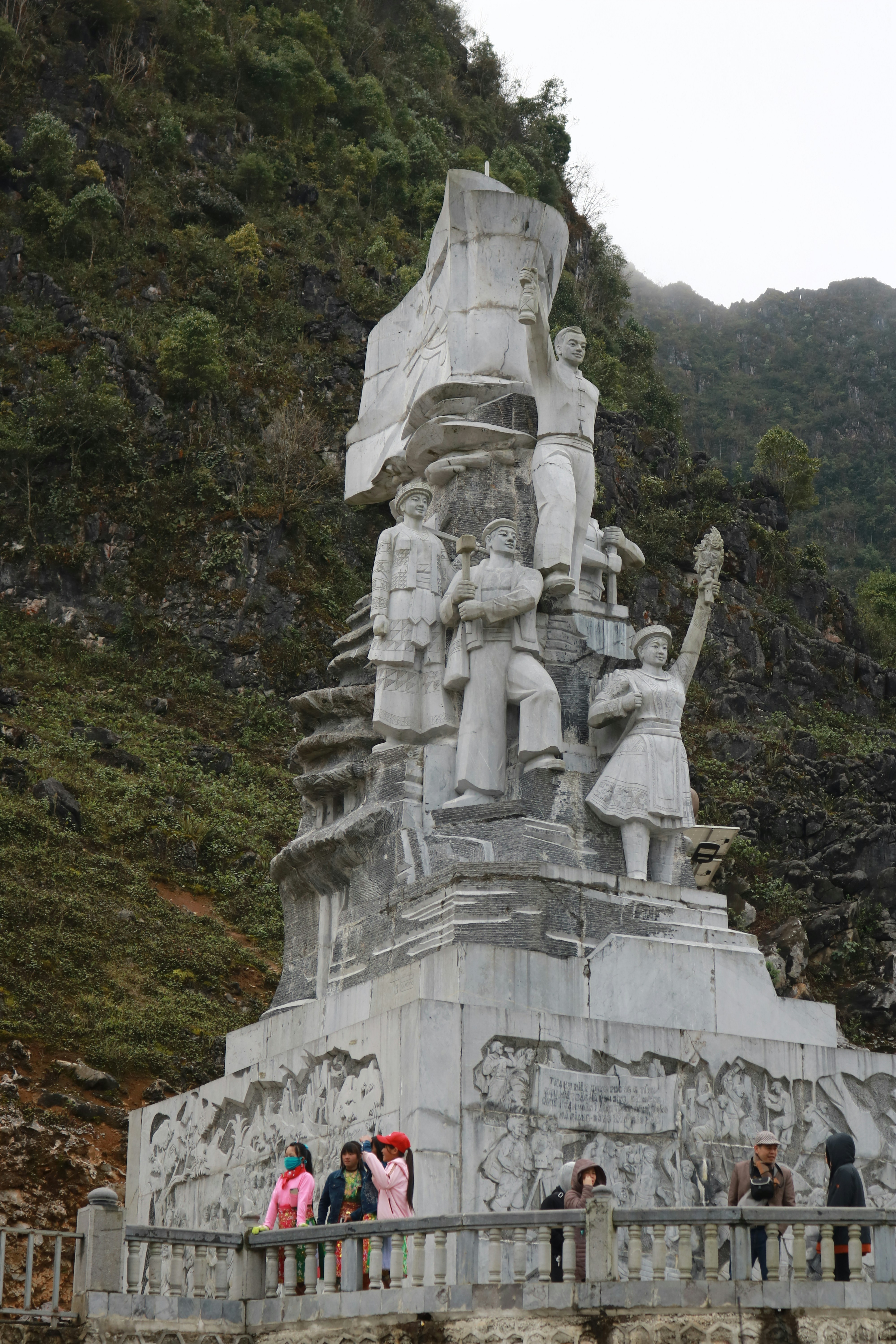 A towering white statue commemorating cultural heritage, featuring figures of workers and historical symbols, set against a lush mountainous backdrop.