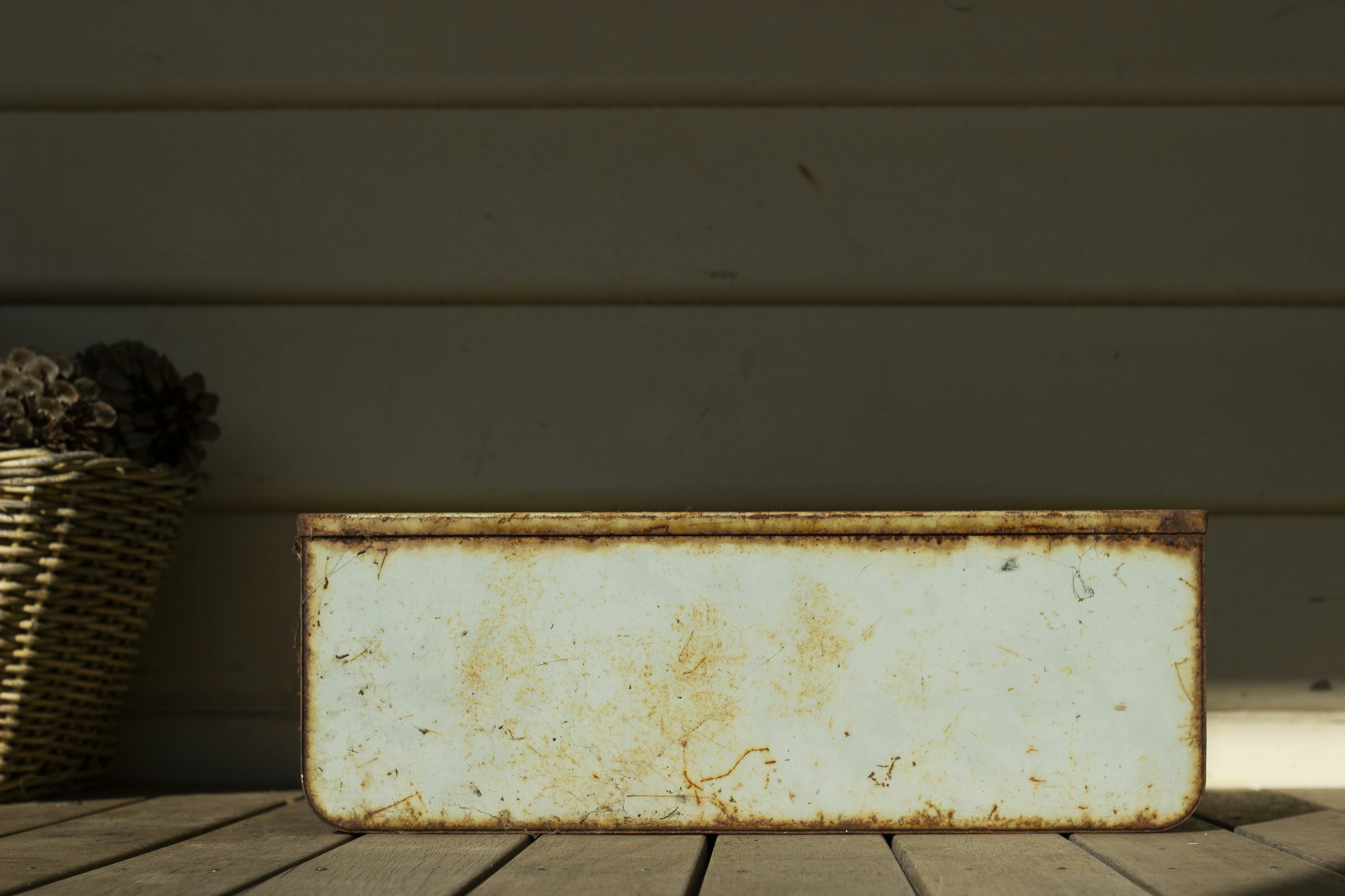 A weathered, light blue container rests on wooden planks, illuminated by soft natural light, with a woven basket and pinecones in the background.