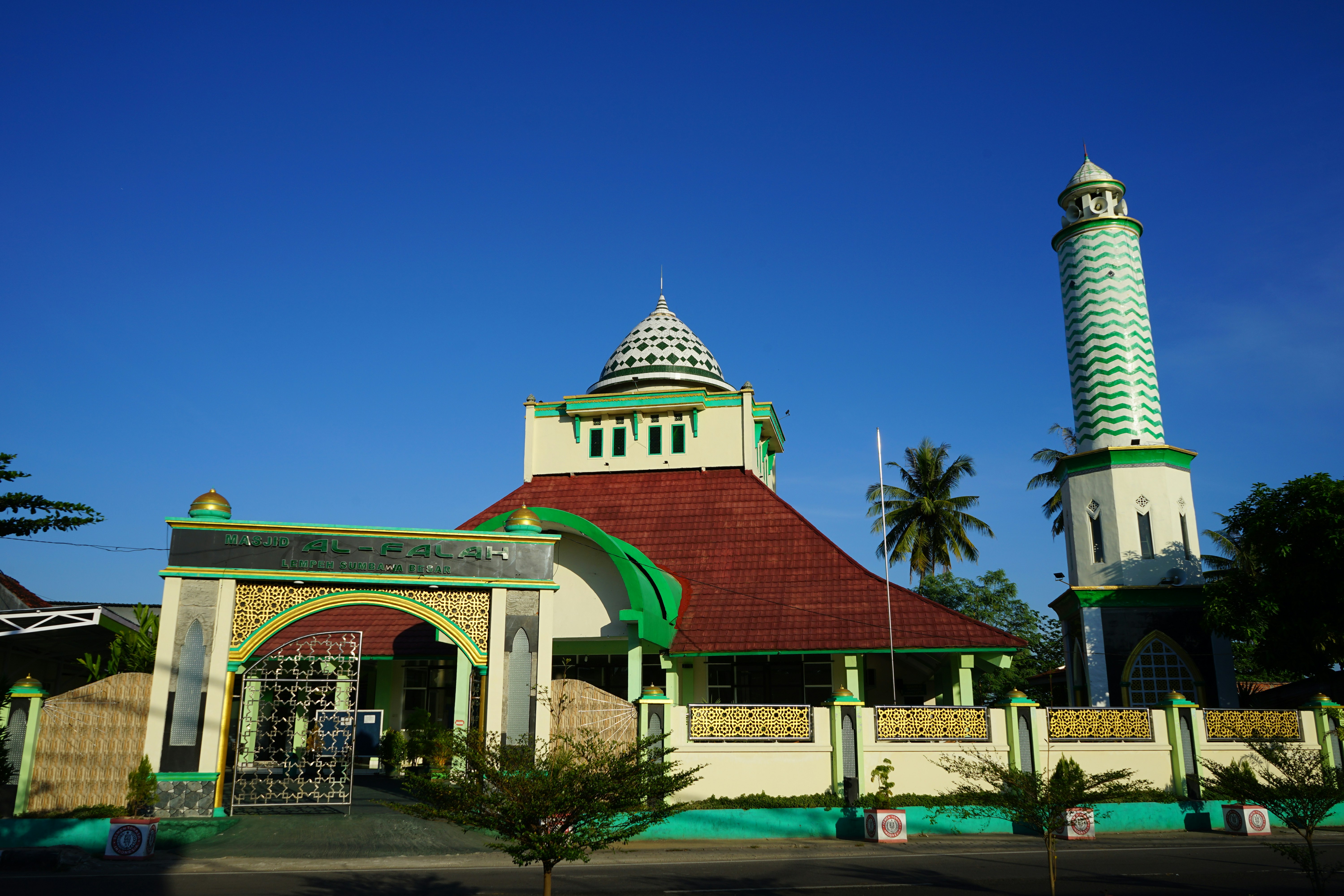 A vibrant mosque showcasing intricate architectural details with a prominent minaret, surrounded by lush greenery and a clear blue sky.
