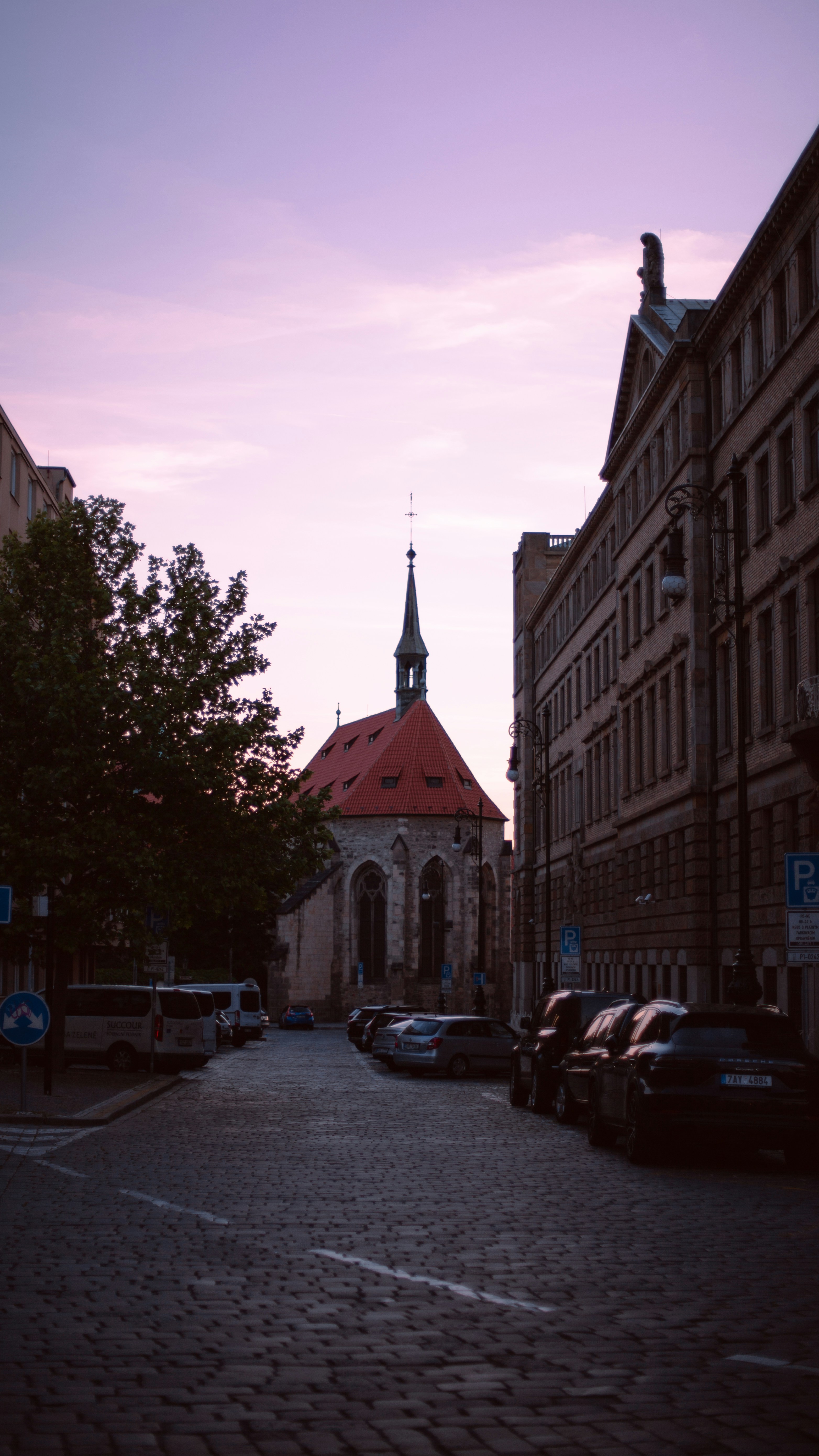 Historic church with a red roof framed by cobblestone street and trees at dusk. Streetlights begin to illuminate the scene.