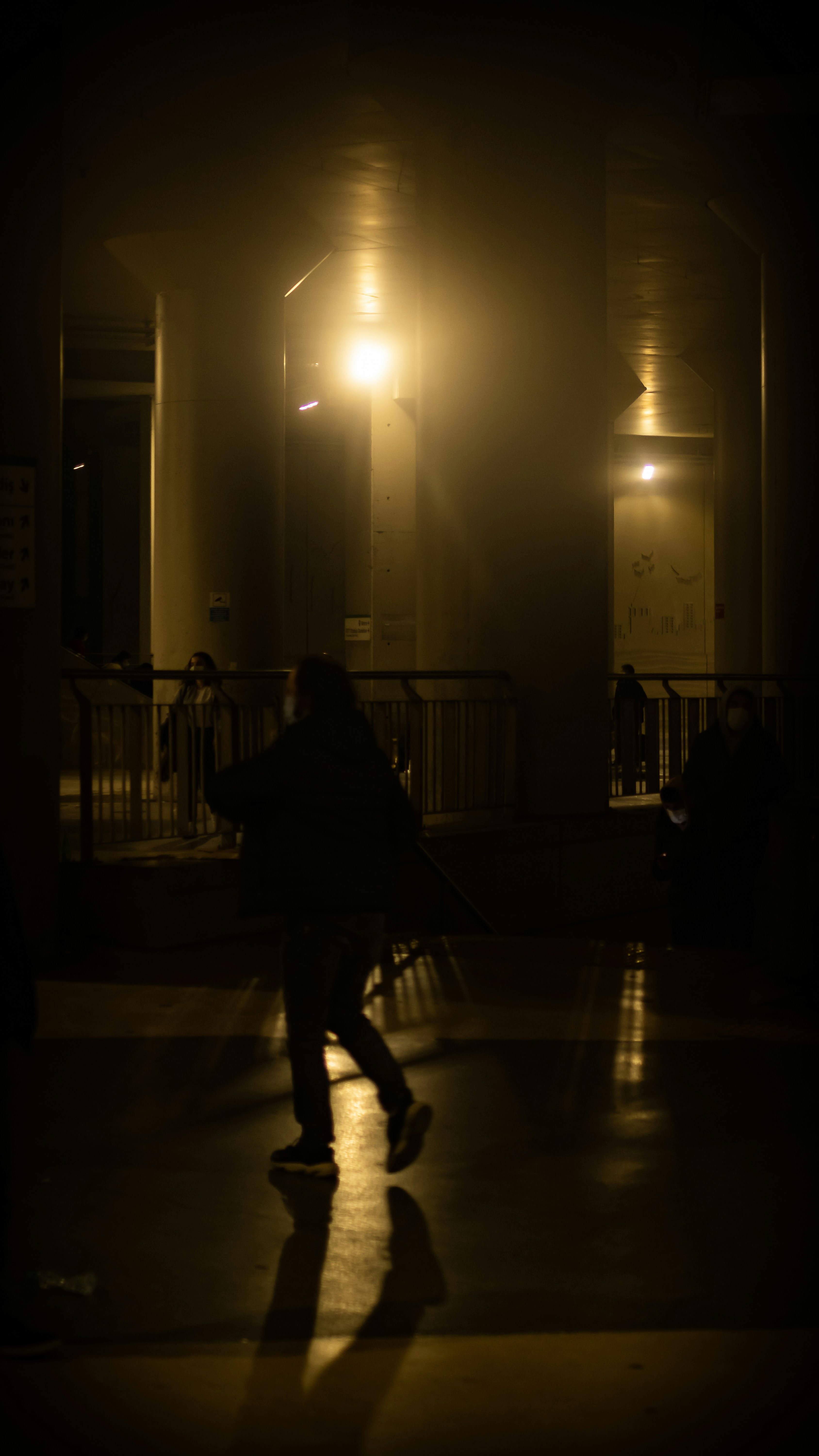 A figure walks purposefully through a dimly lit urban space, illuminated by soft overhead lights. The contrast of shadows and light creates an atmospheric scene.