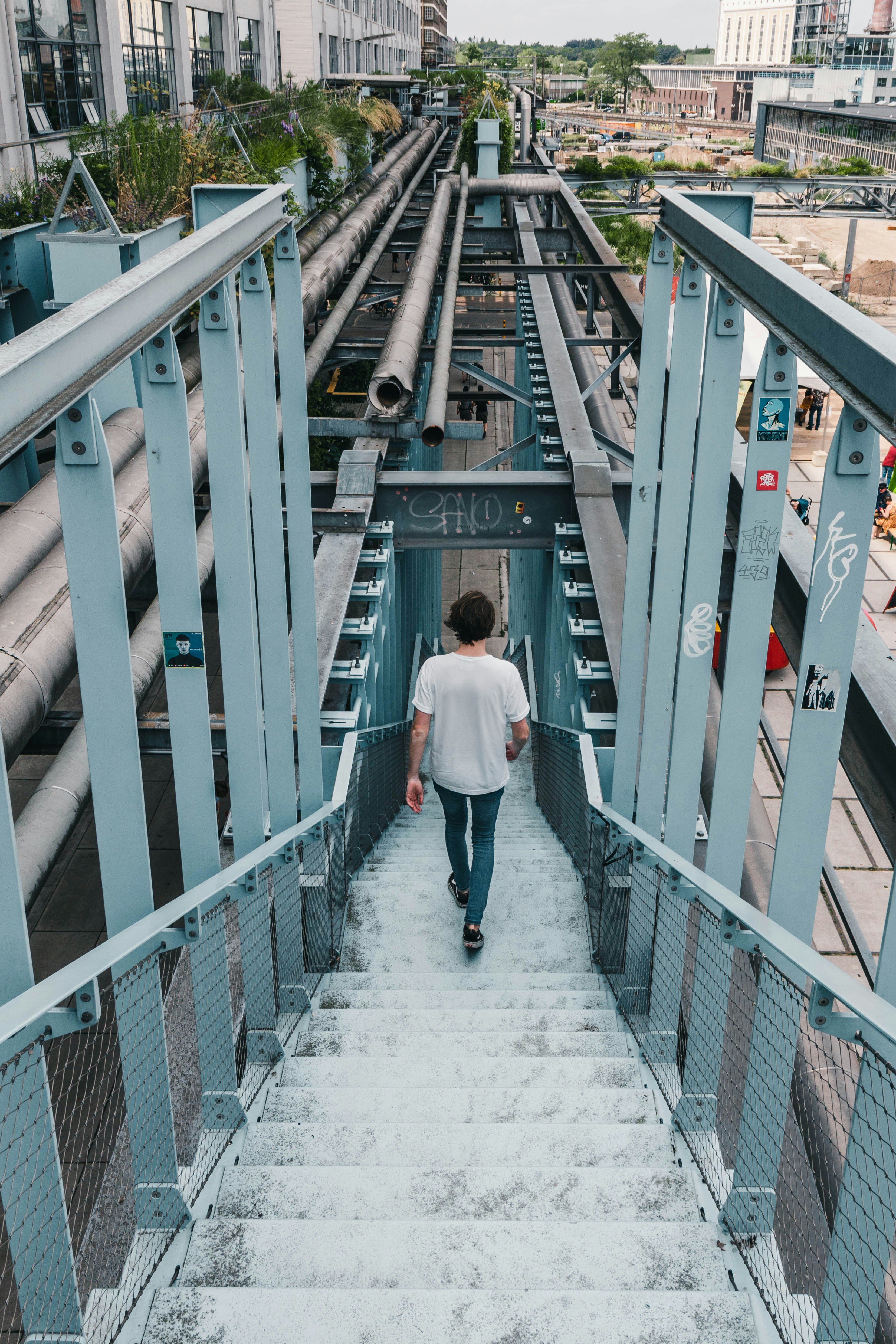 A person descends a staircase surrounded by industrial pipes, leading to a vibrant urban landscape. The scene captures the intersection of man-made structures and urban exploration.