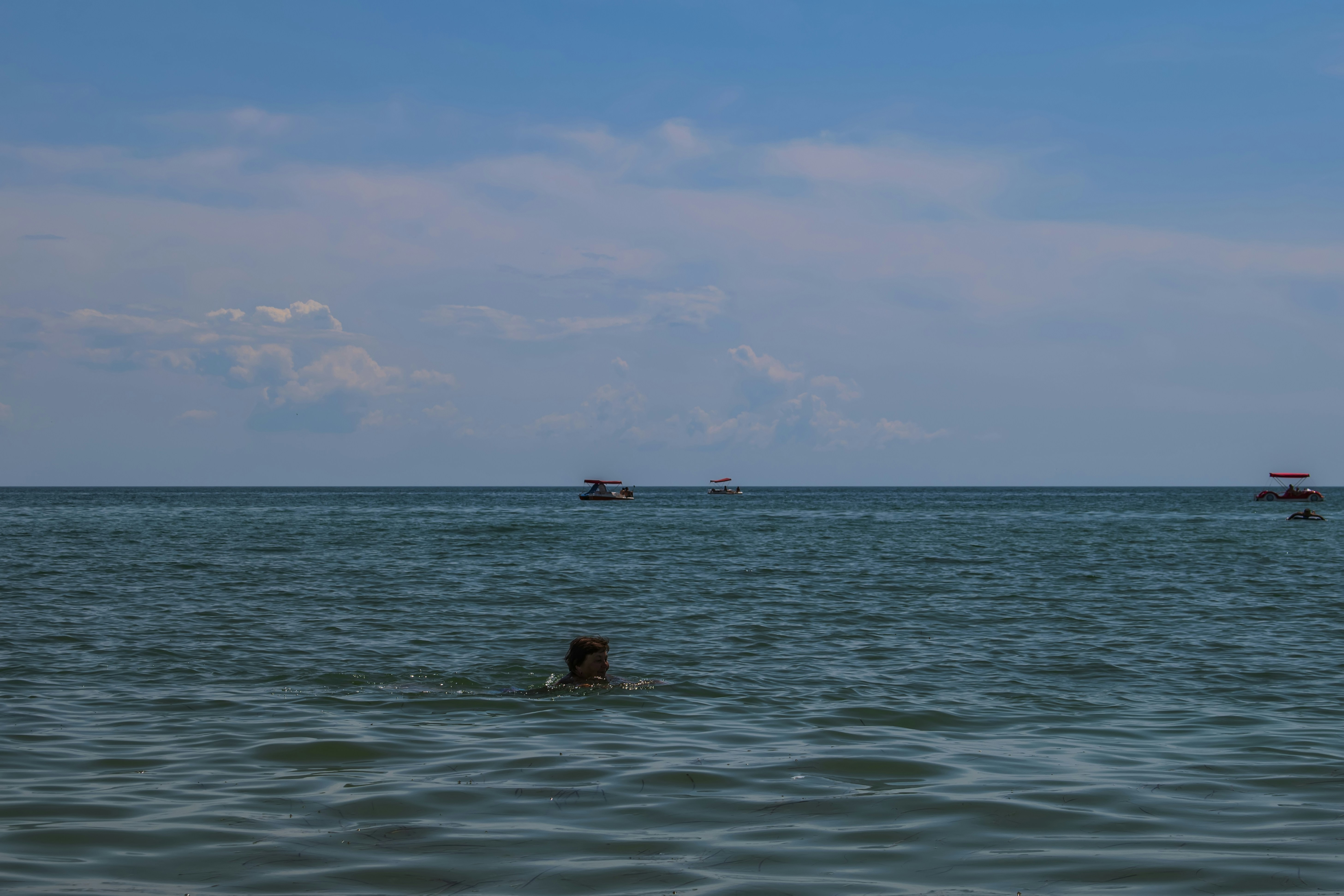 A swimmer glides through tranquil waters under a clear sky, with distant boats dotting the horizon. The scene conveys a sense of peaceful leisure.
