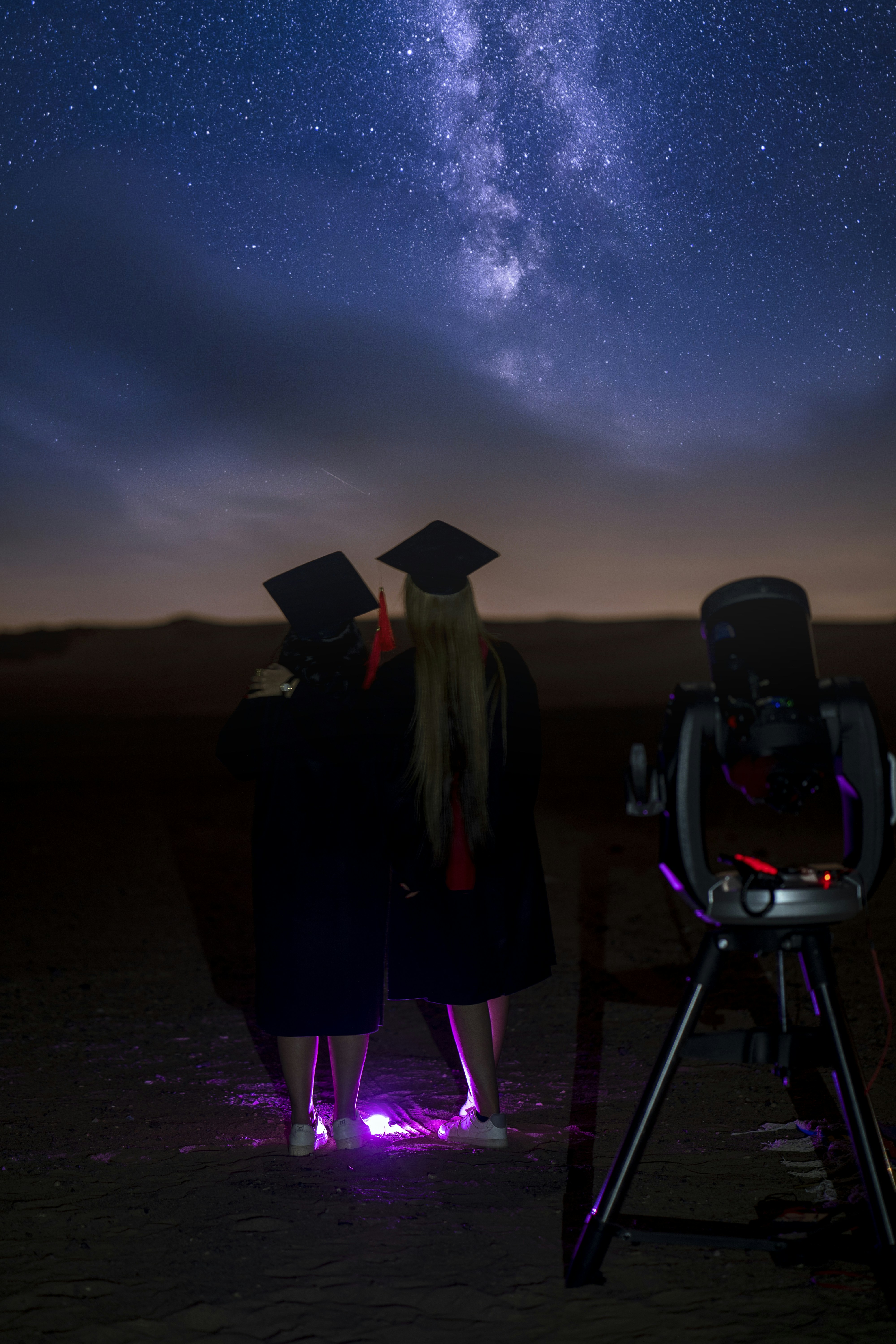 Two graduates in caps and gowns stand together, gazing at the Milky Way above, illuminated by soft ground lights. A telescope nearby hints at a night of exploration.