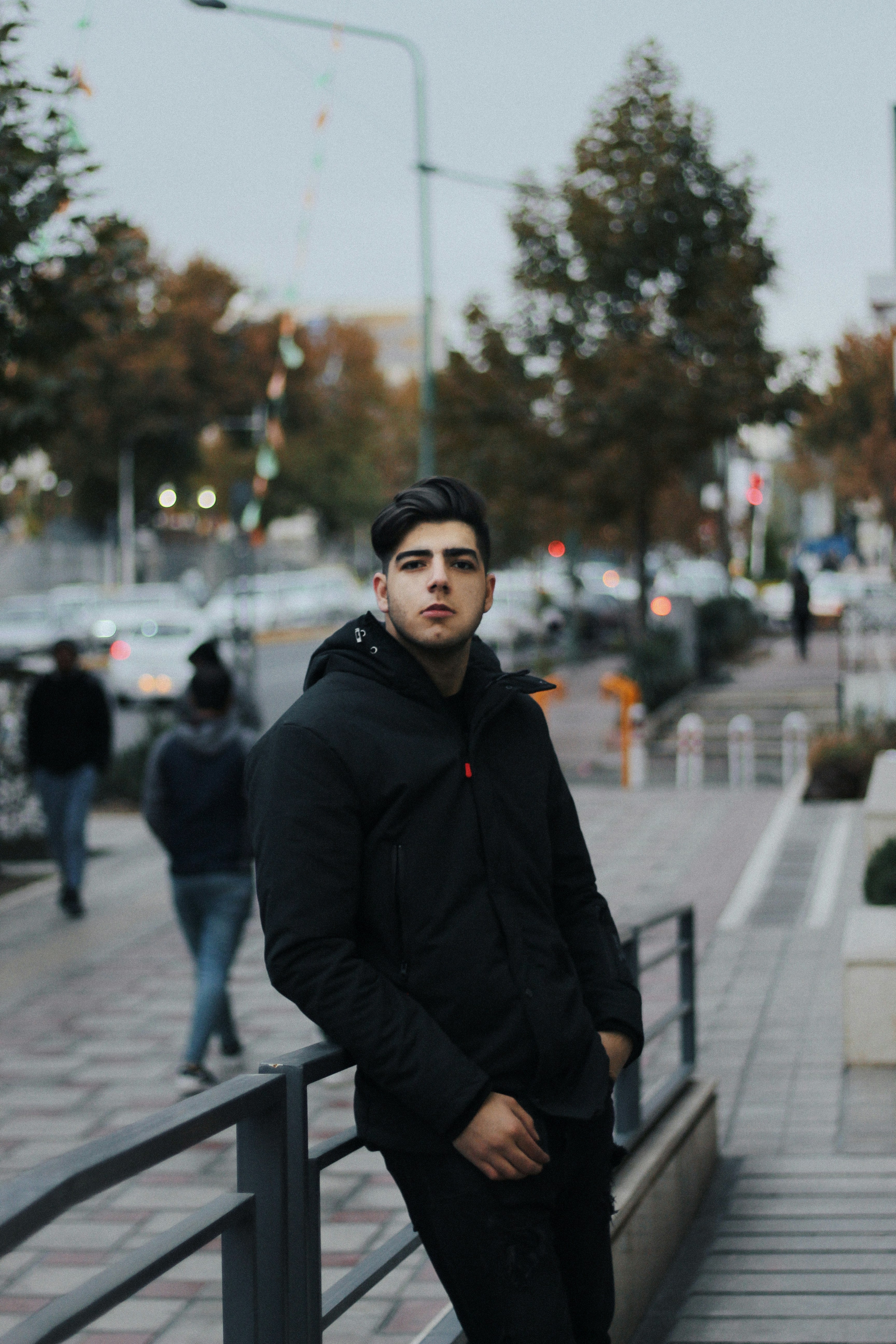 Young man in a black jacket leaning against a railing in a bustling city street, surrounded by blurred pedestrians and autumn foliage.