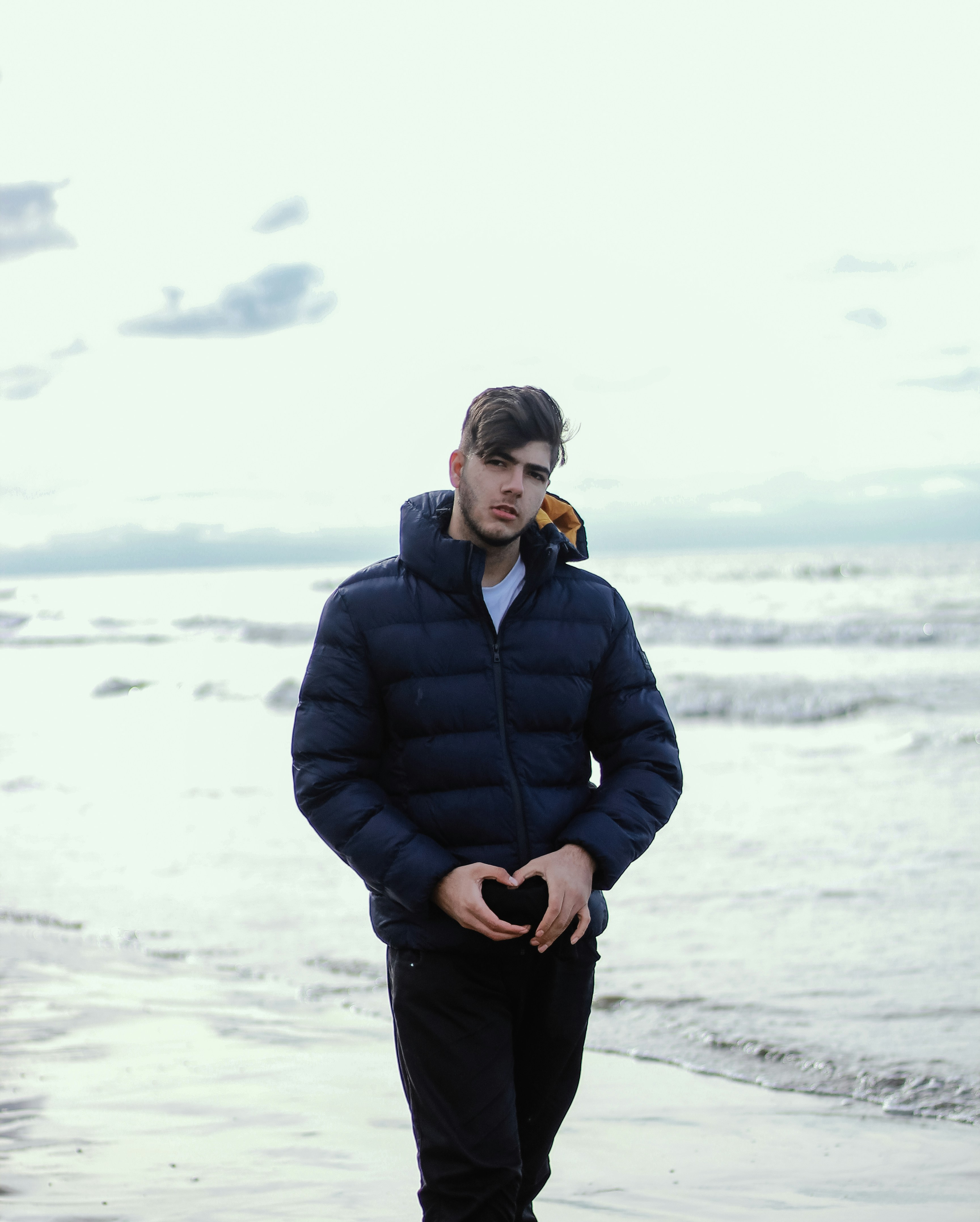 Young man forming a heart shape with his hands while standing on a beach, with gentle waves lapping at the shore. The cloudy sky creates a serene atmosphere.