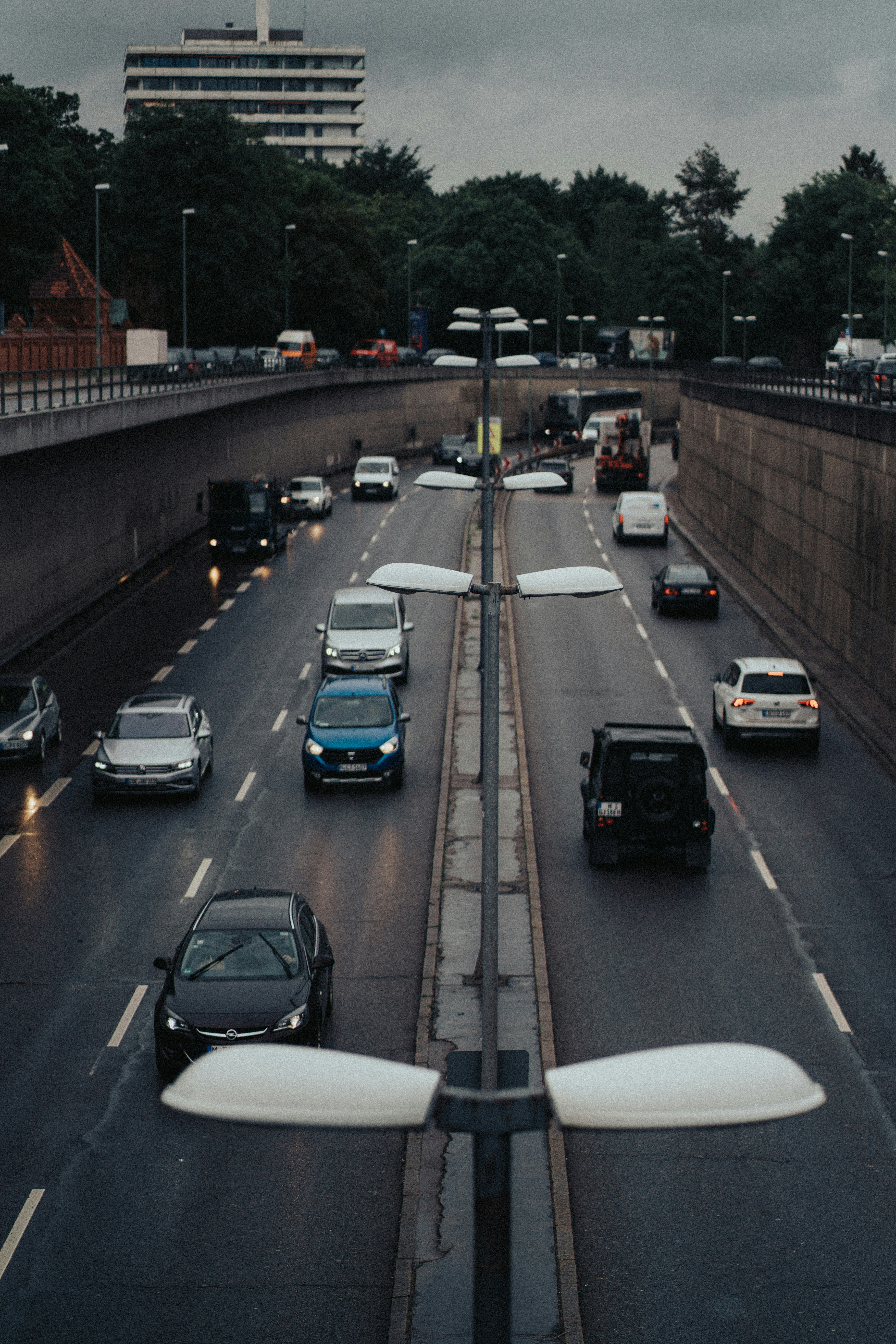 Traffic navigating a sunken roadway under overcast skies, with streetlights illuminating the scene. The urban landscape features a mix of vehicles and greenery in the background.
