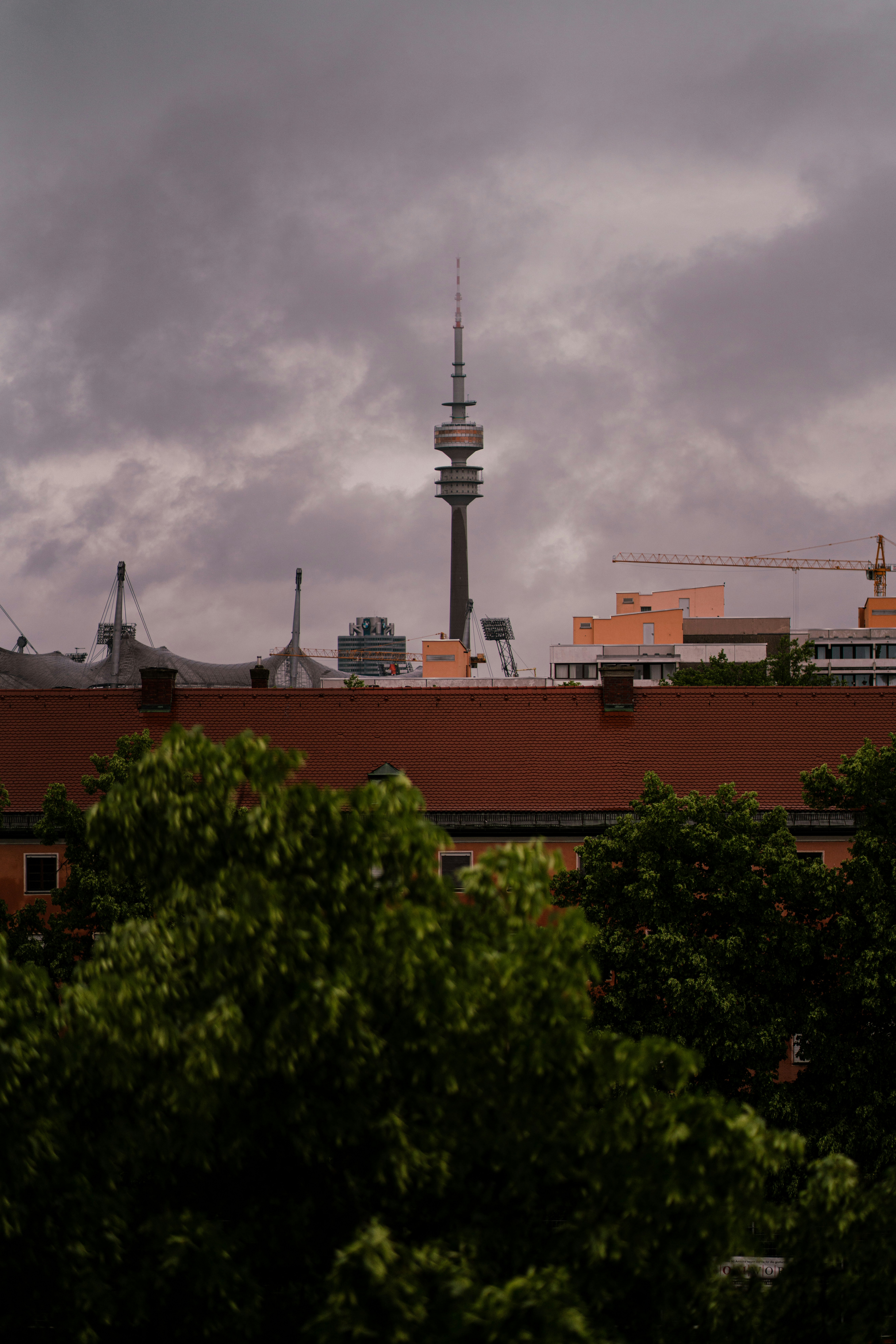 Telecommunication tower rises above rooftops, framed by lush greenery and an overcast sky. The scene captures a blend of urban architecture and nature.