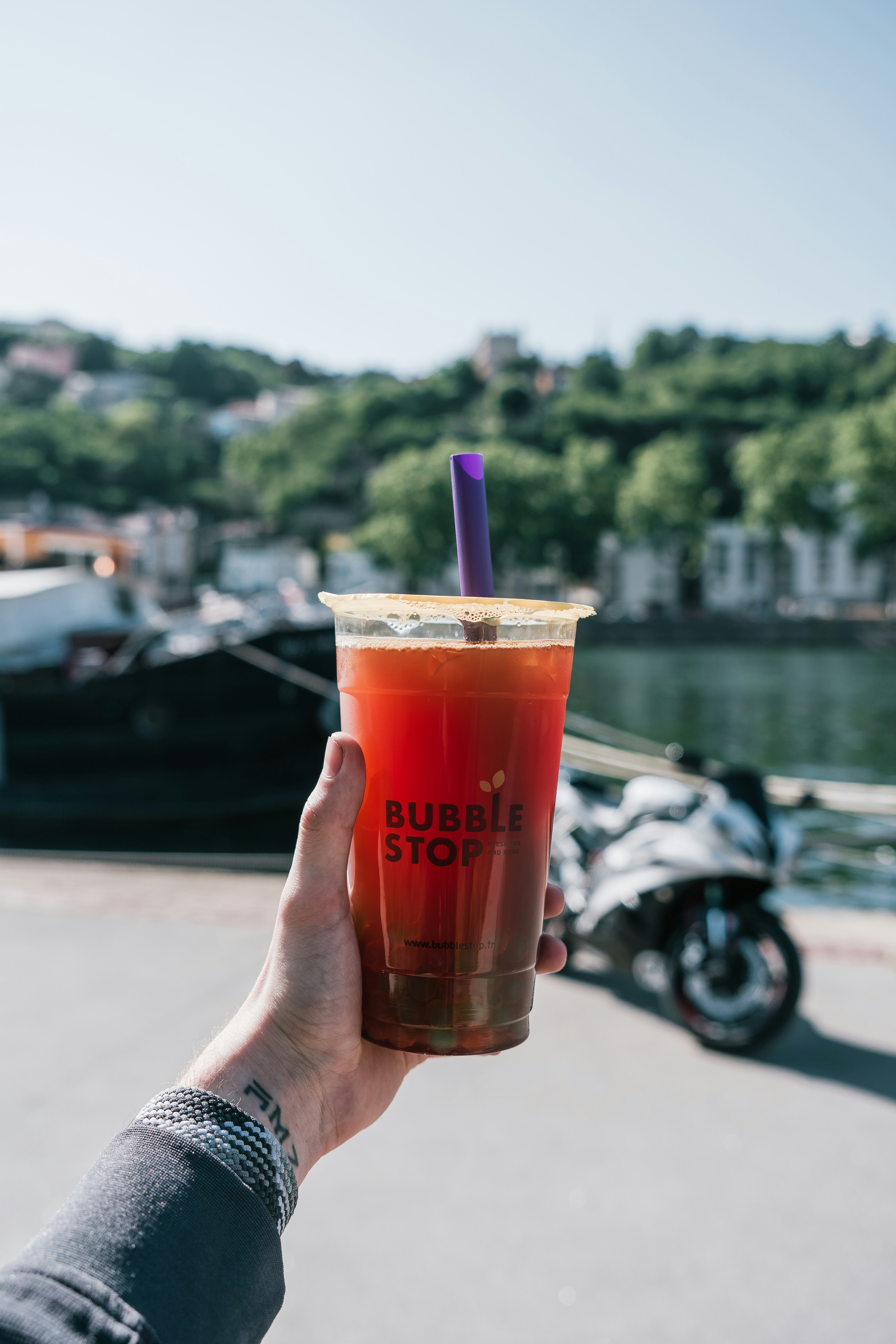 Hand holding a transparent cup of bubble tea with a purple straw, set against a backdrop of lush greenery and a serene waterfront.