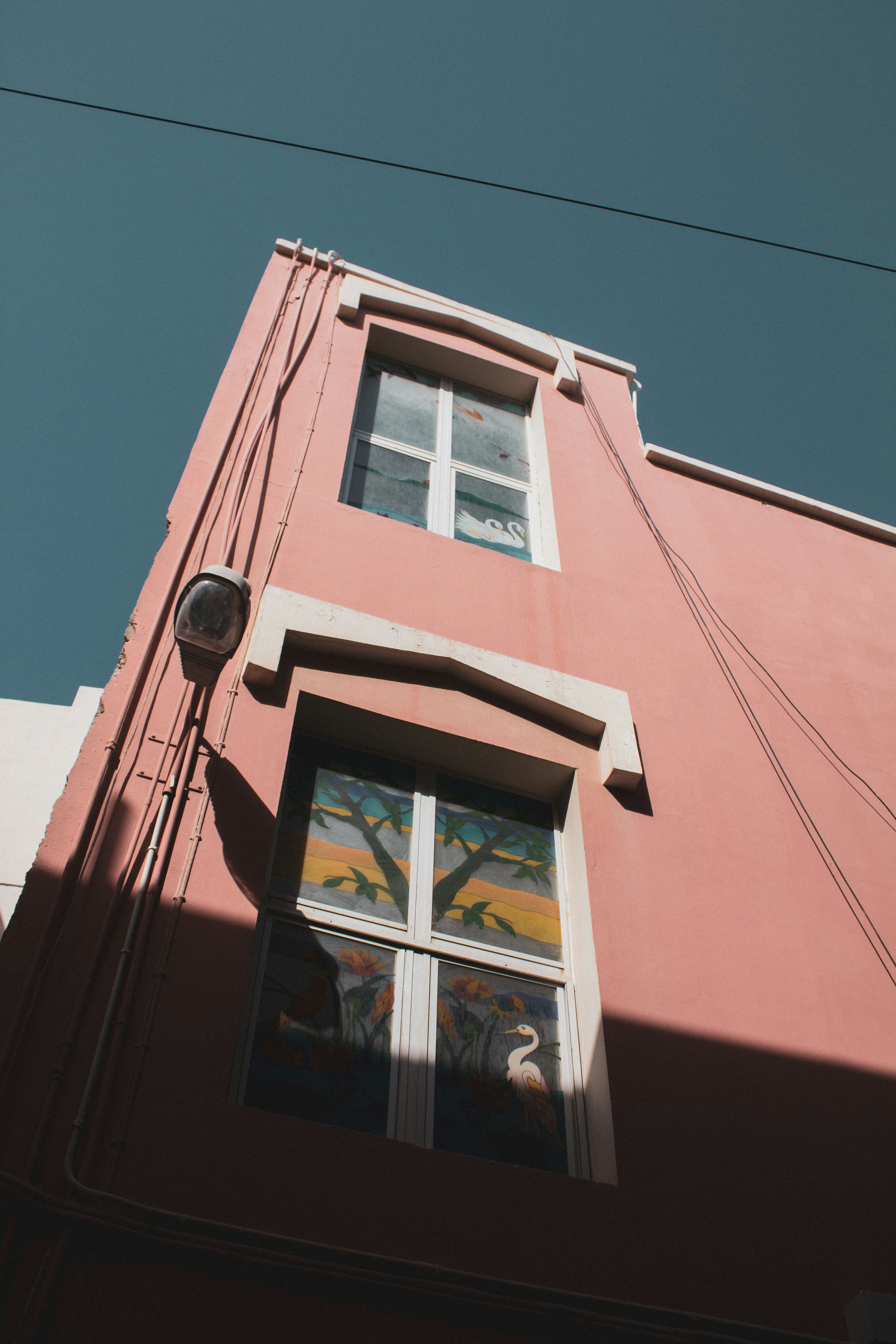 Vibrant mural visible through a window on a pink building, contrasted by shadows and a clear blue sky.