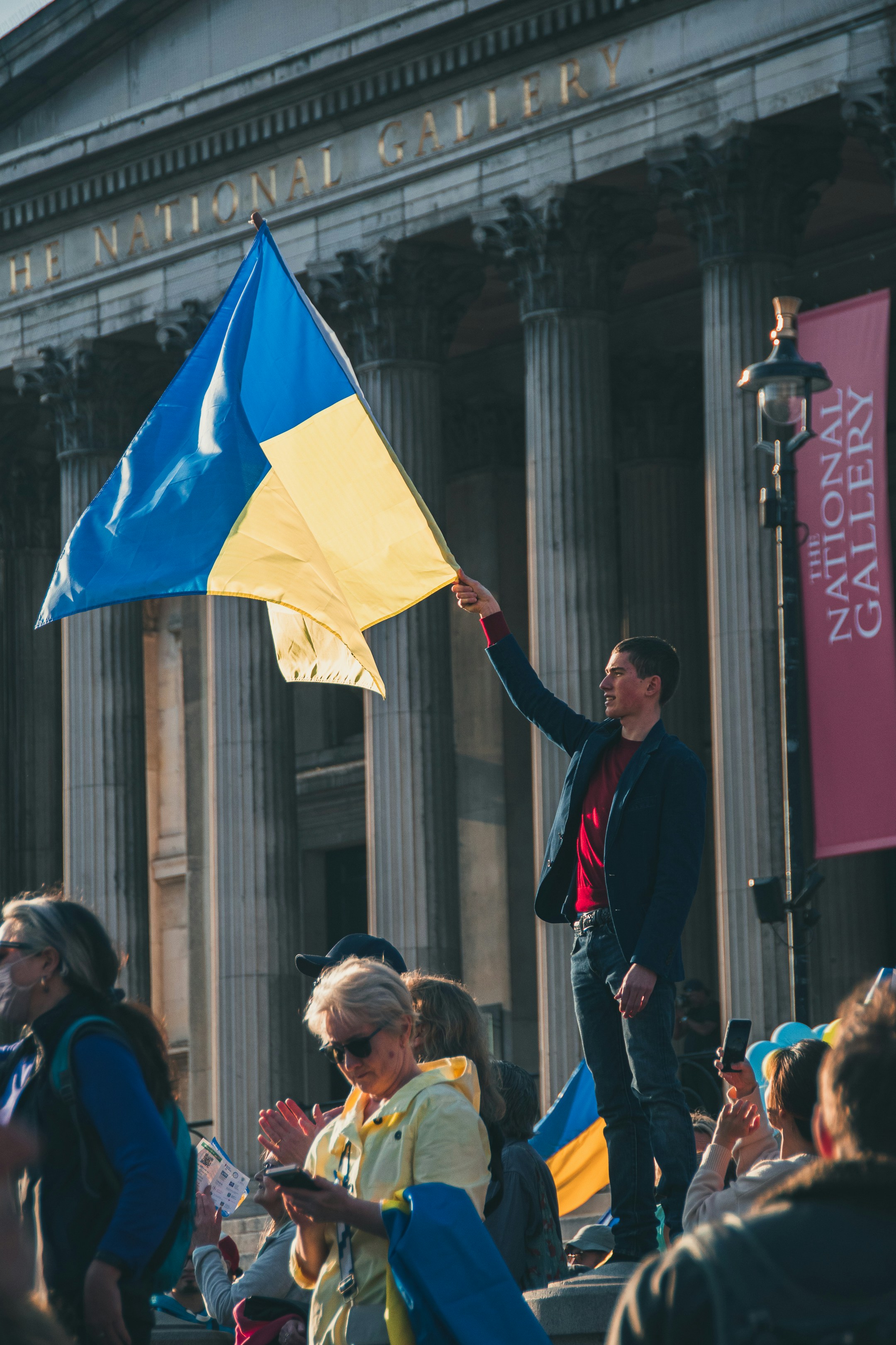 Man proudly waves the Ukrainian flag during a gathering at The National Gallery, surrounded by a diverse crowd showing support.