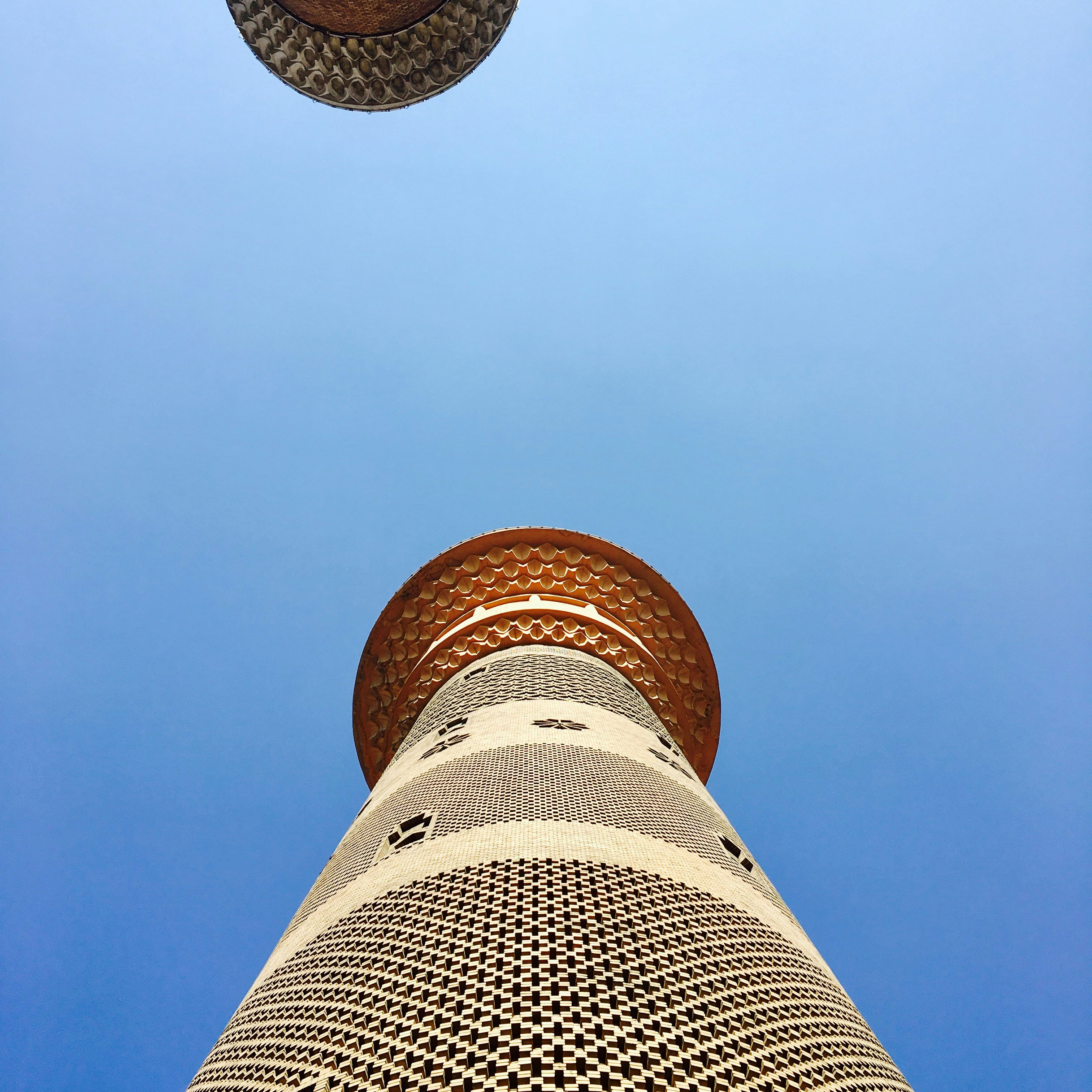 Intricate patterned column rising towards a clear blue sky, showcasing artistic craftsmanship and design. A decorative cap rests above, enhancing the vertical perspective.