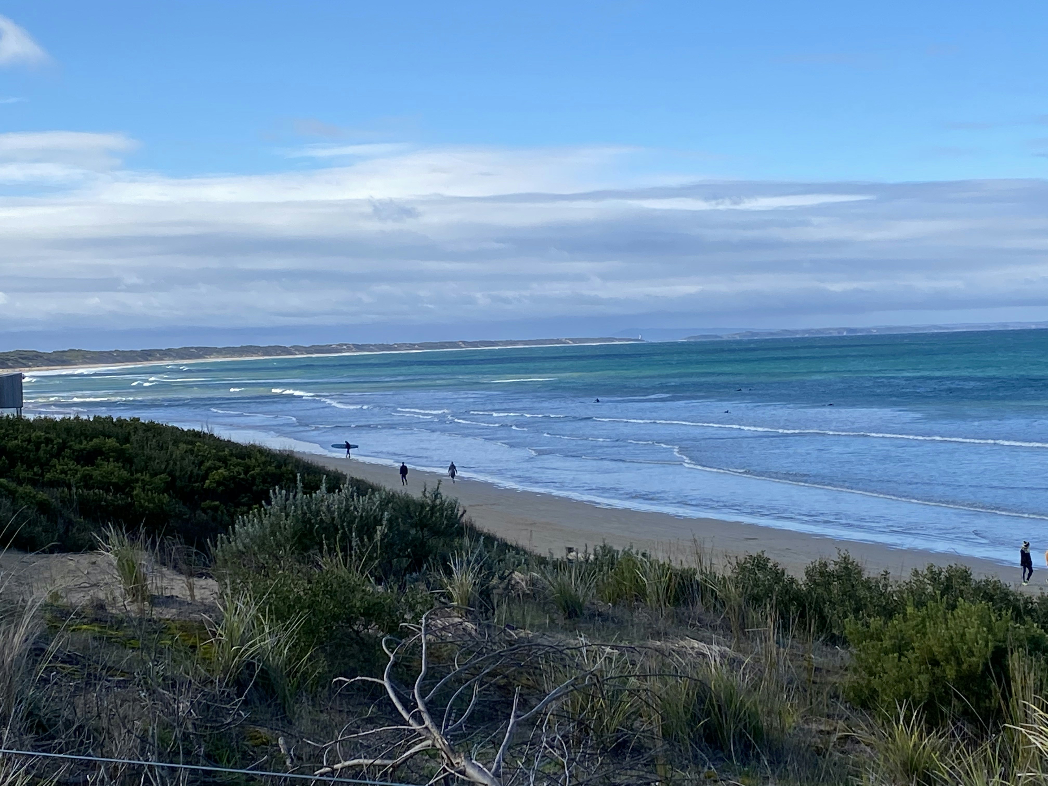 Waves gently lap against the sandy shore, with silhouettes of beachgoers enjoying the tranquil seaside atmosphere. Lush greenery frames the shoreline.