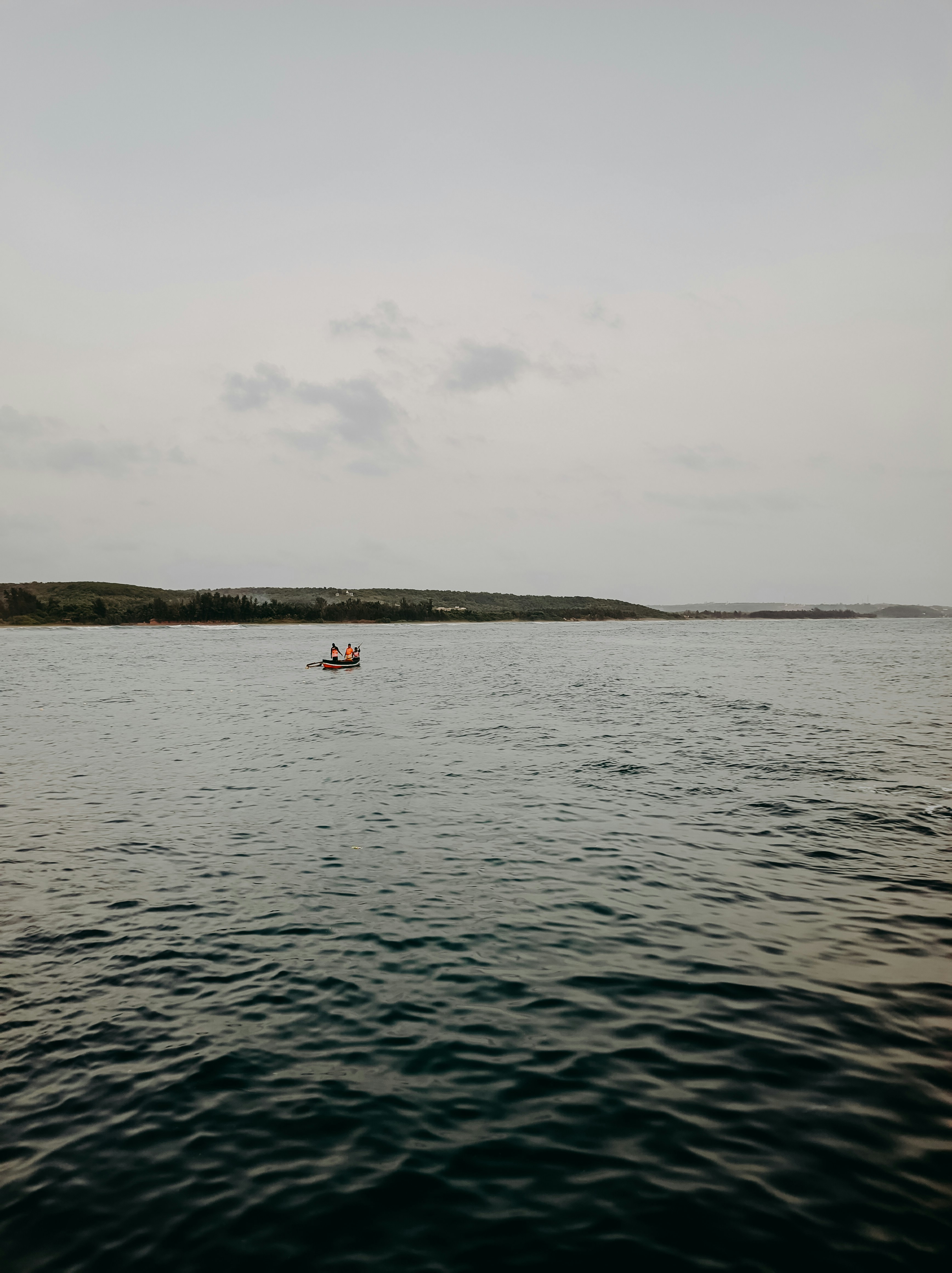 Calm seascape photograph featuring a small boat with two people on tranquil waters. A distant coastline lies beneath a pale, overcast sky.