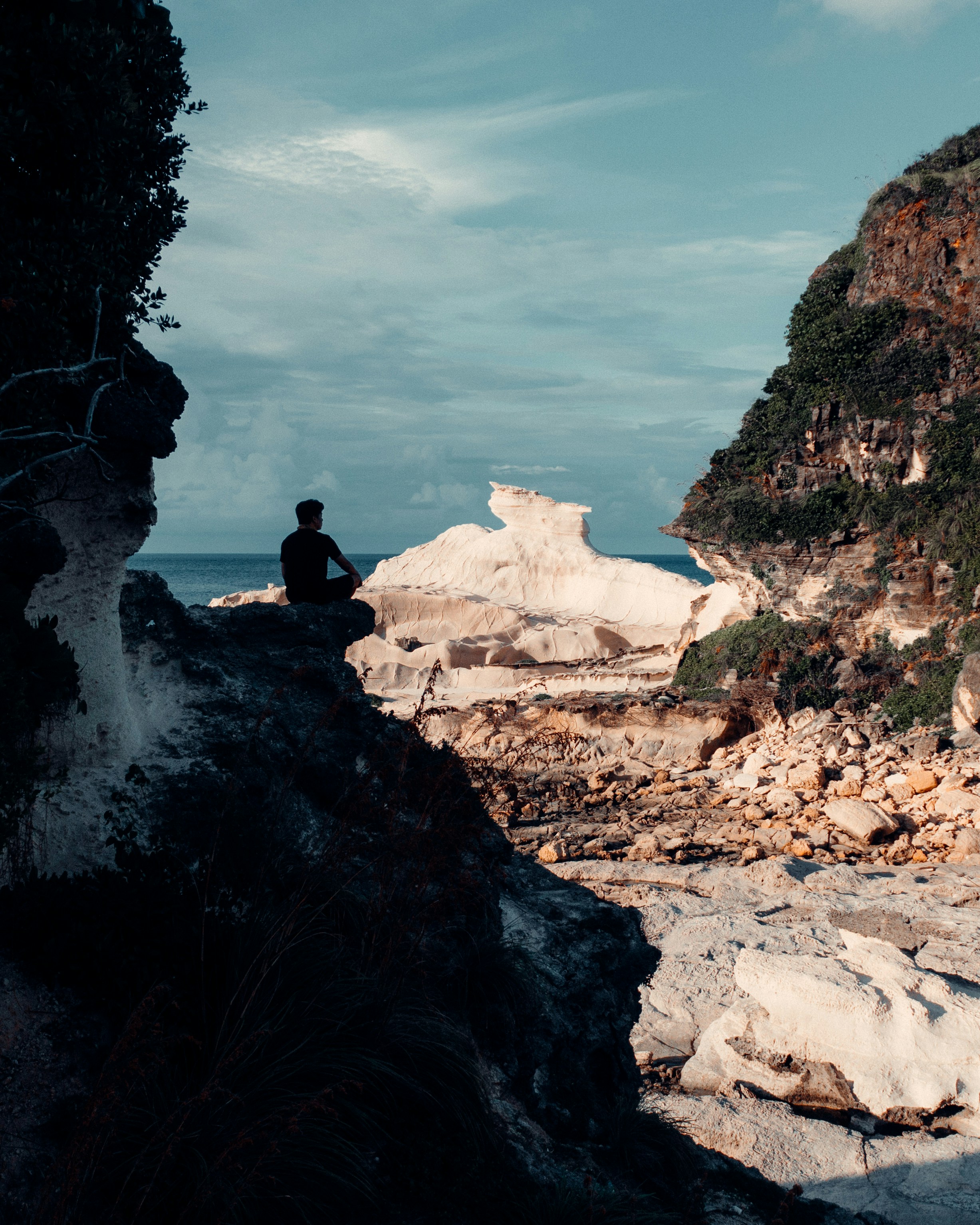A solitary figure sits on a rocky outcrop, gazing at the unique white formations along the coastline, where land meets sea under a serene sky.