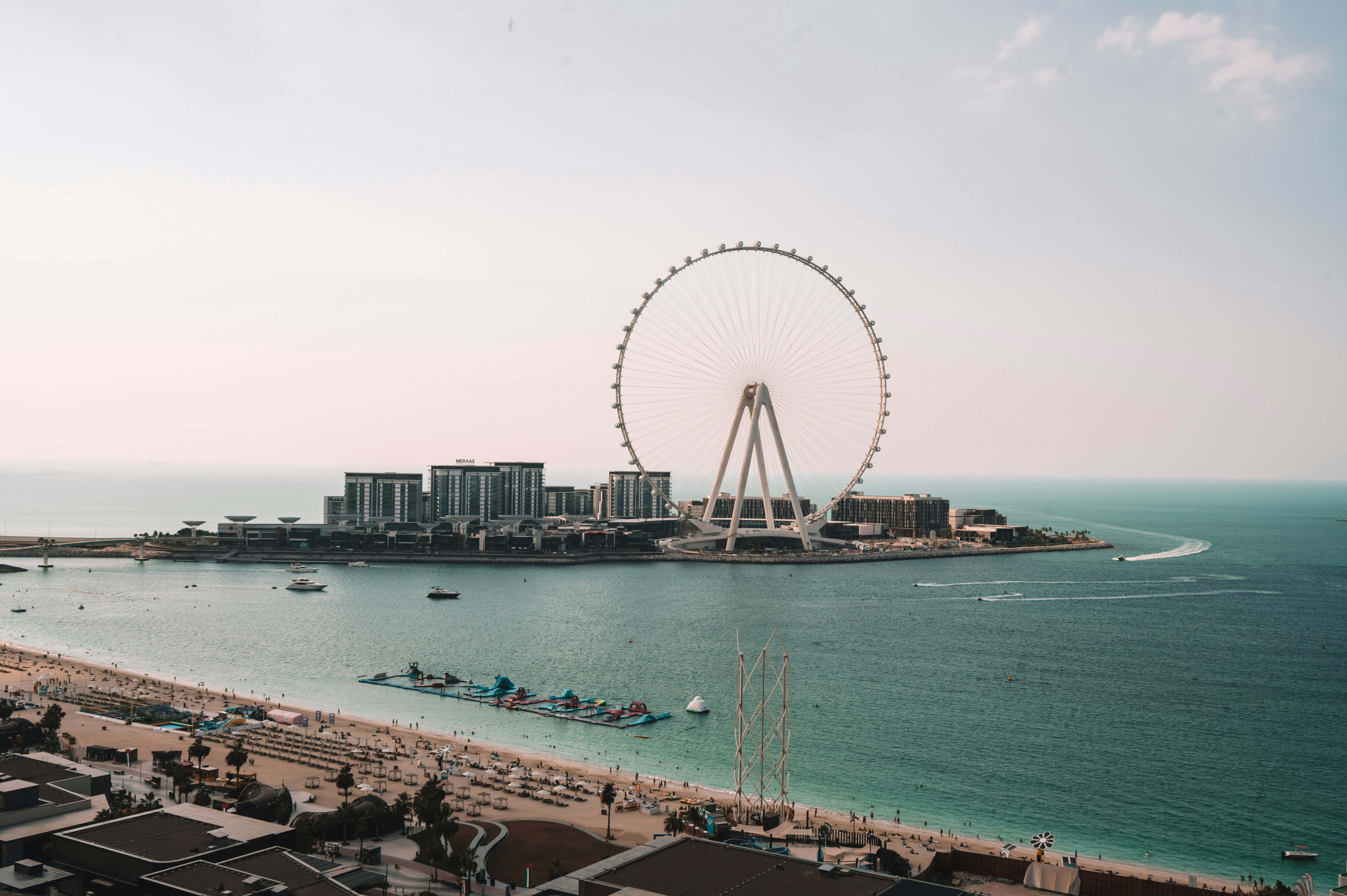 A panoramic view of a coastal landscape featuring a large Ferris wheel and contemporary buildings along the shore, with tranquil waters reflecting the sky.