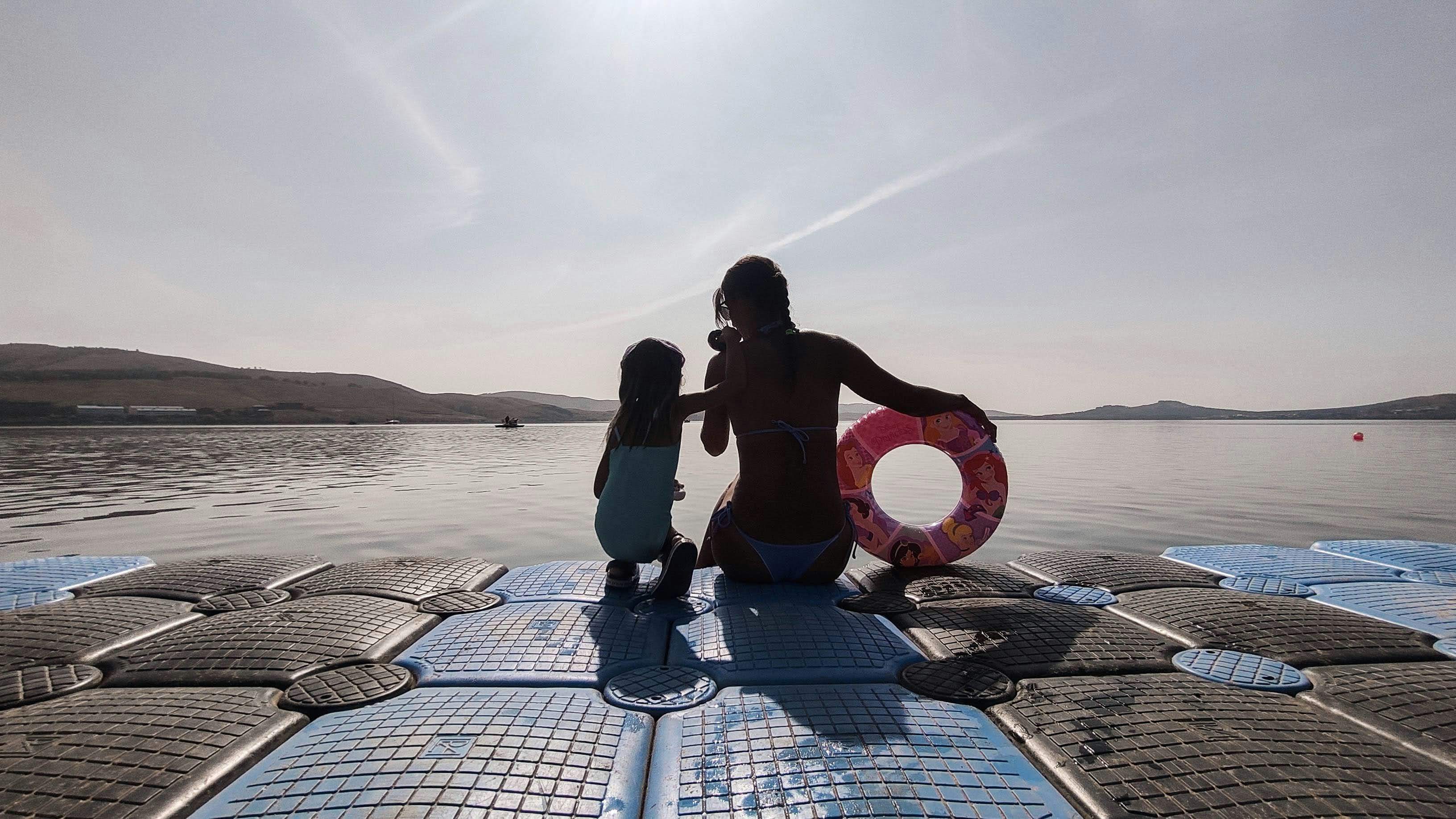 Silhouetted figures of a woman and child sitting on a floating platform, enjoying a serene moment by the water. A colorful float ring rests beside them.