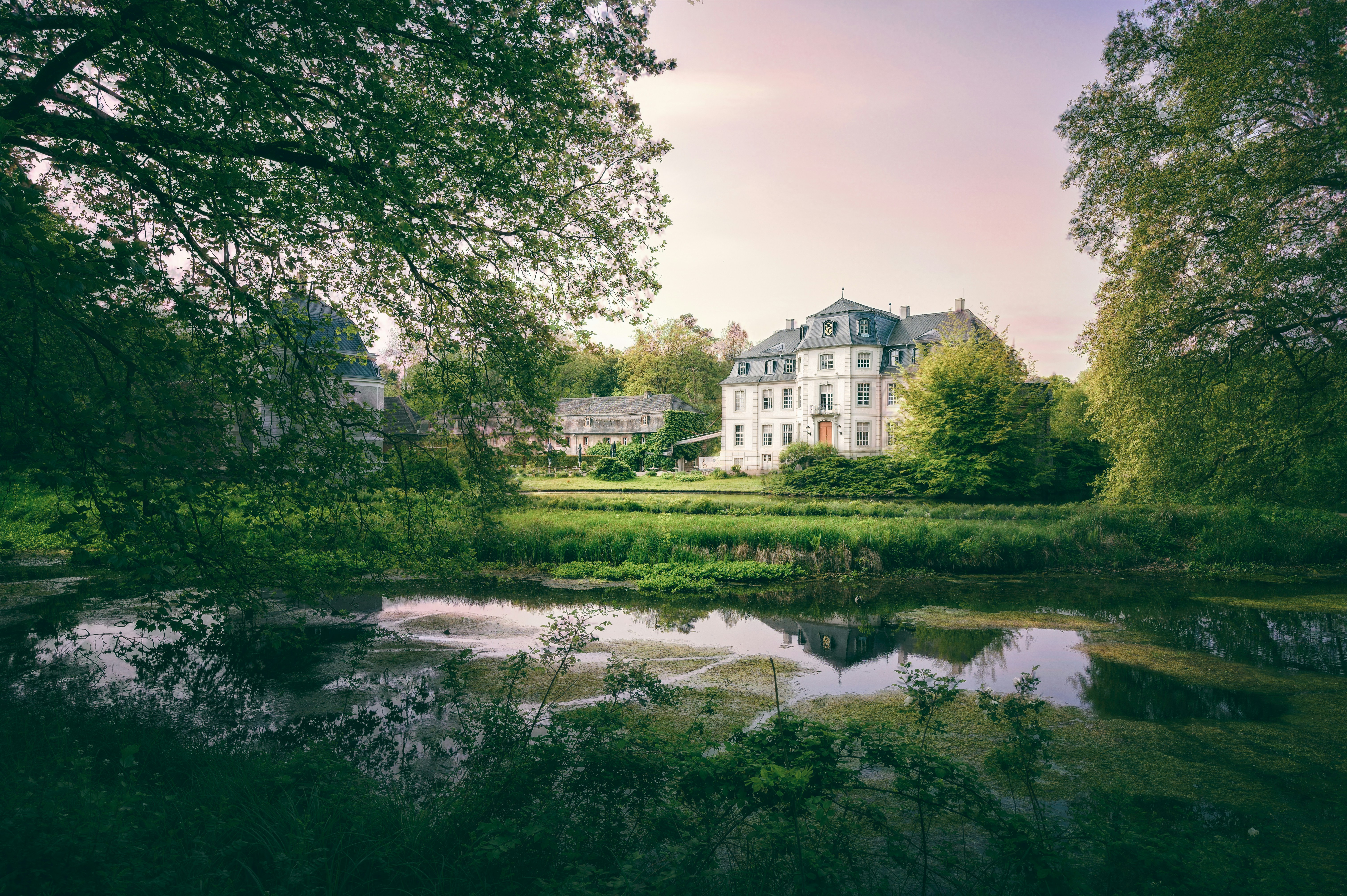 Elegant manor reflected in a serene pond, framed by lush greenery under a pastel sky.
