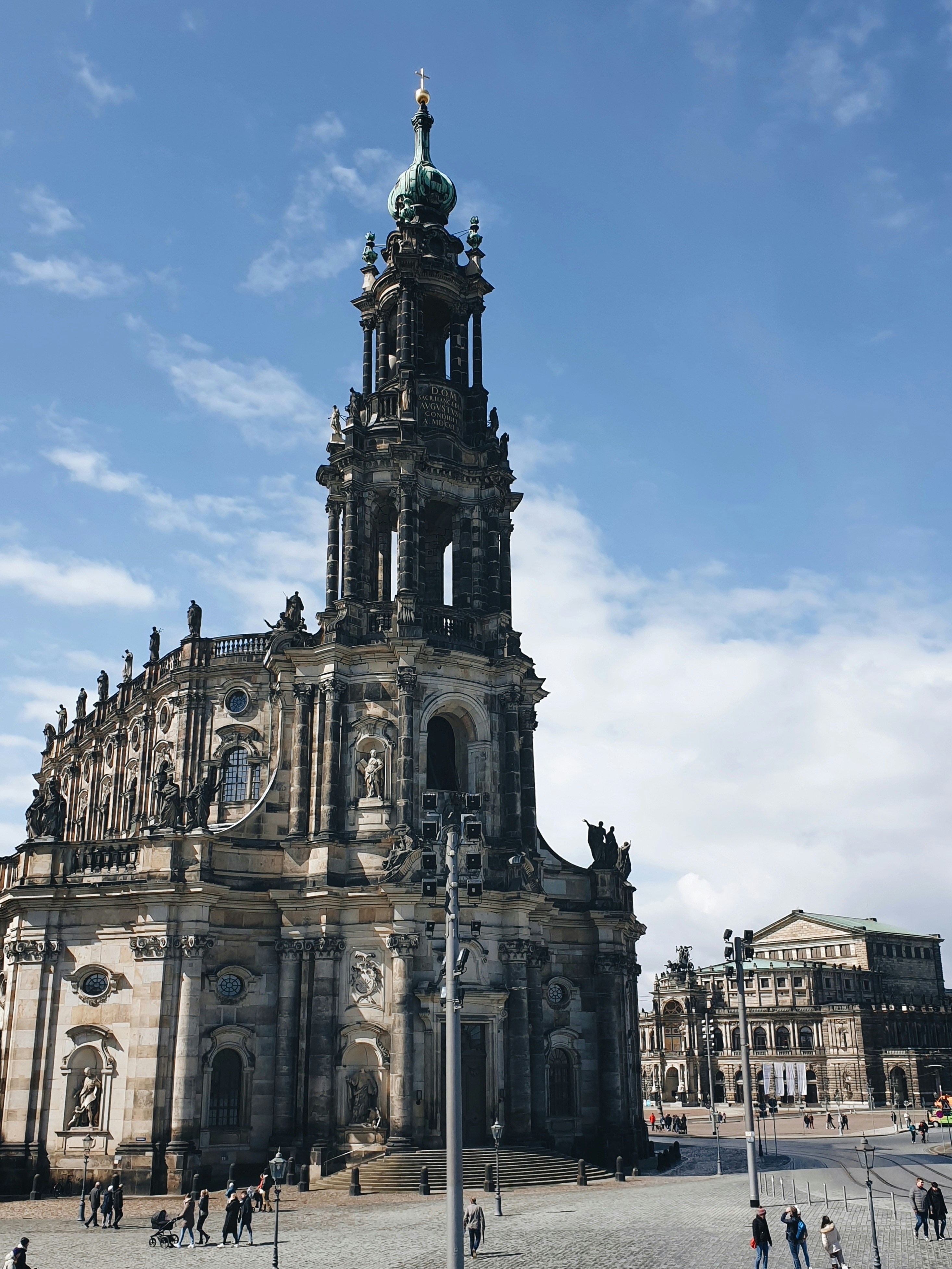 Baroque church facade adorned with intricate sculptures and a green dome, set against a bright blue sky. Tourists explore the plaza below.