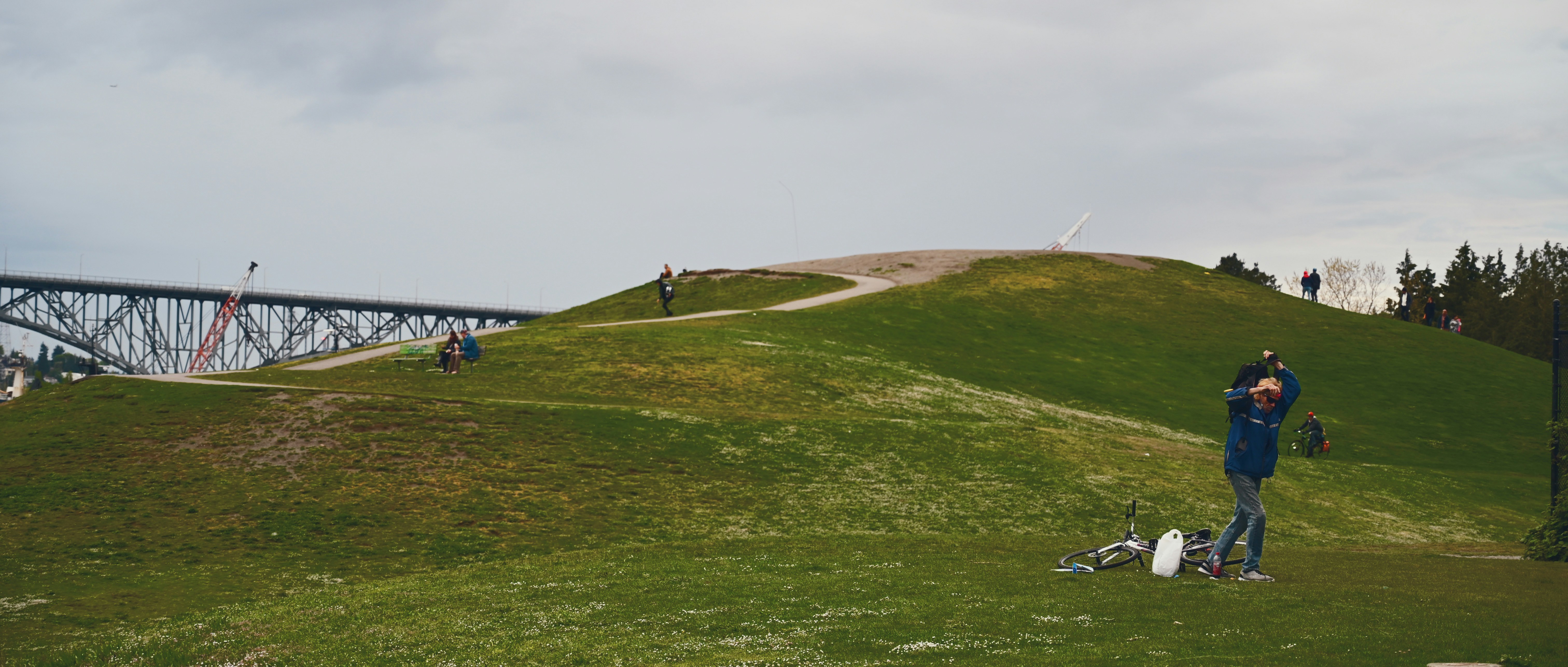 A group of people enjoying leisure activities on a grassy hill, with a bridge visible in the background. The scene captures a relaxed outdoor atmosphere.
