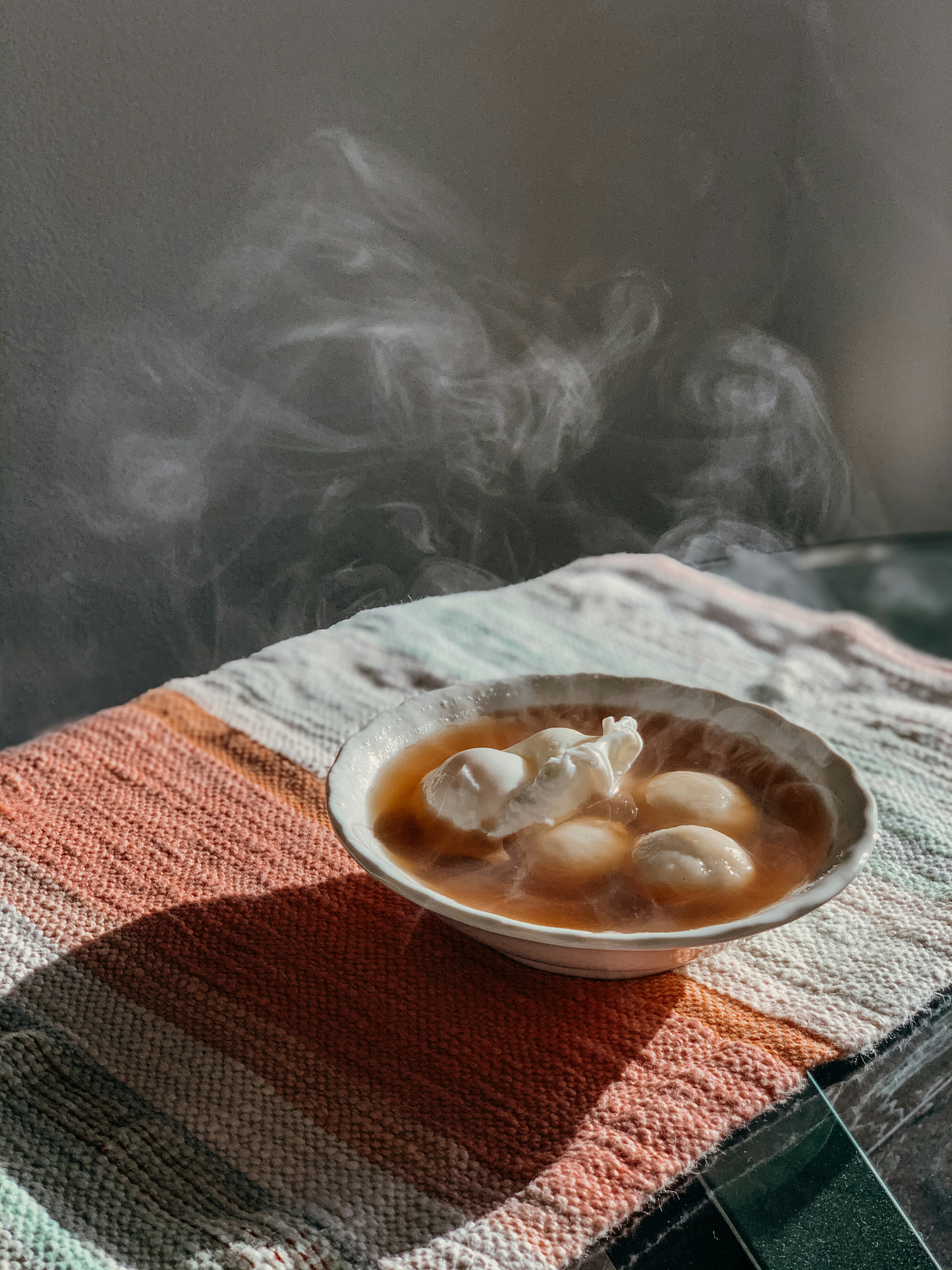 A bowl of poached eggs resting in a warm, savory broth, surrounded by wisps of steam, set against a textured, colorful cloth.