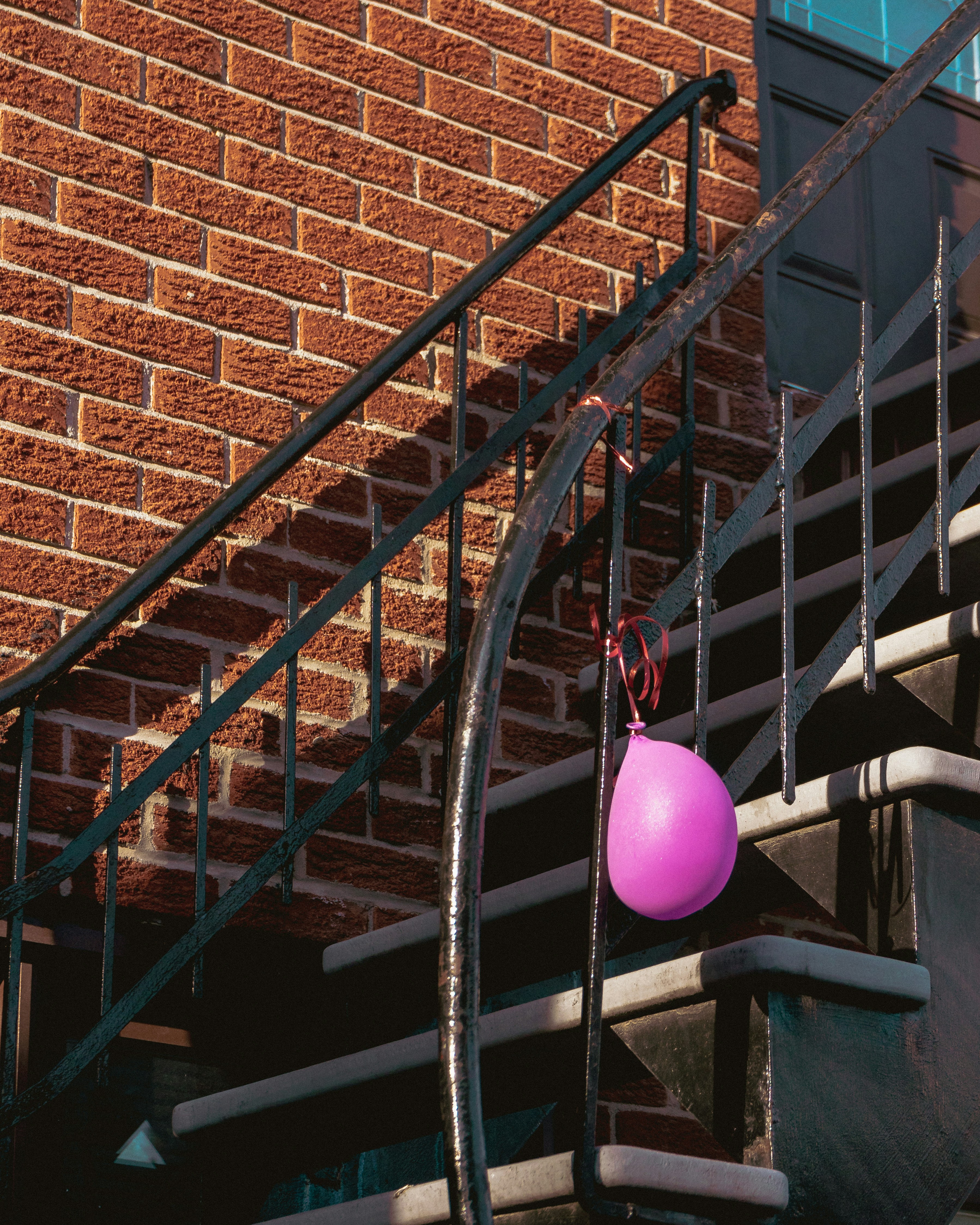 Pink Balloon Dangling Along a Curved Fire Escape Against Red Brick