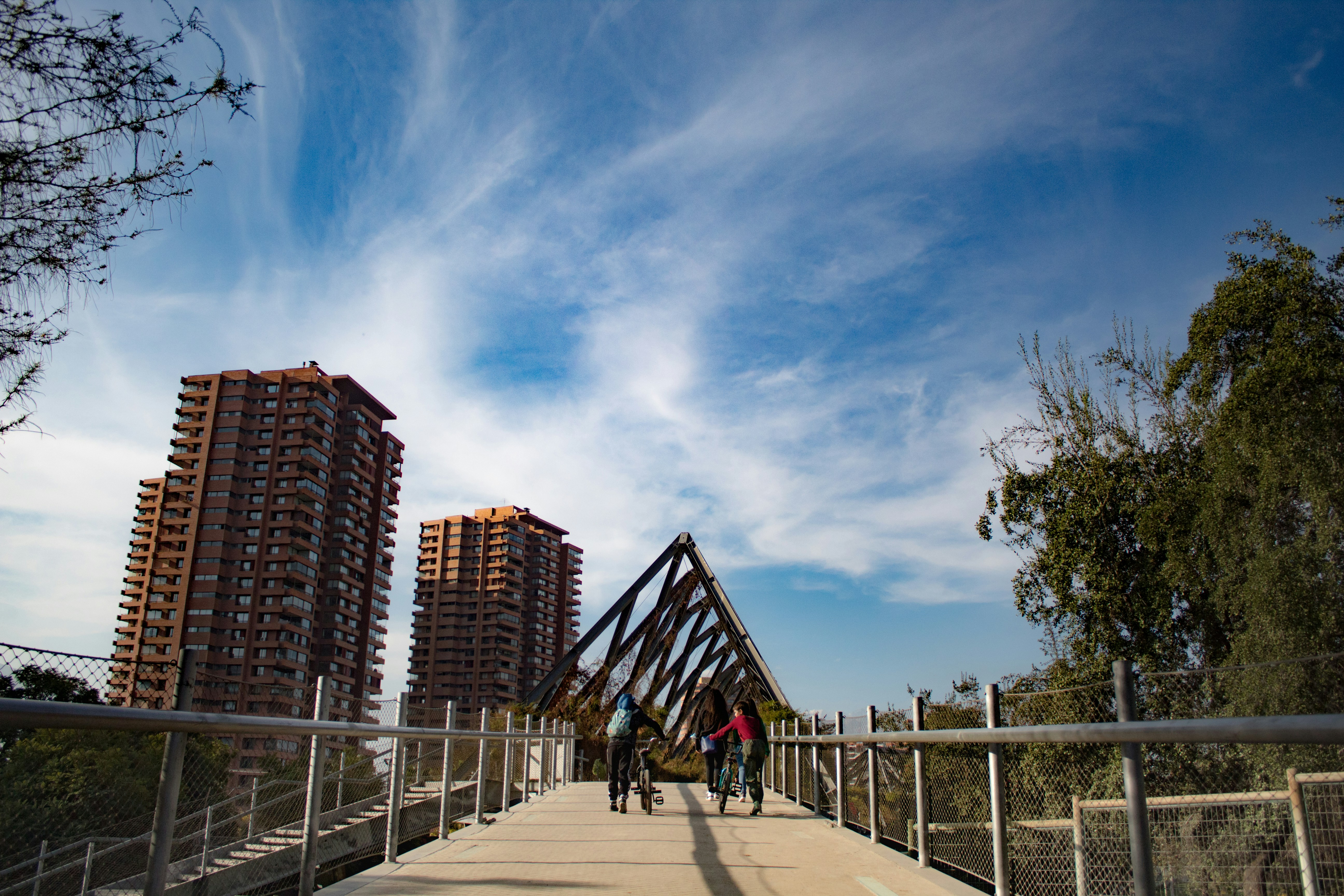 Pedestrians walk on a bridge with geometric railings, flanked by tall buildings and trees under a vivid blue sky.