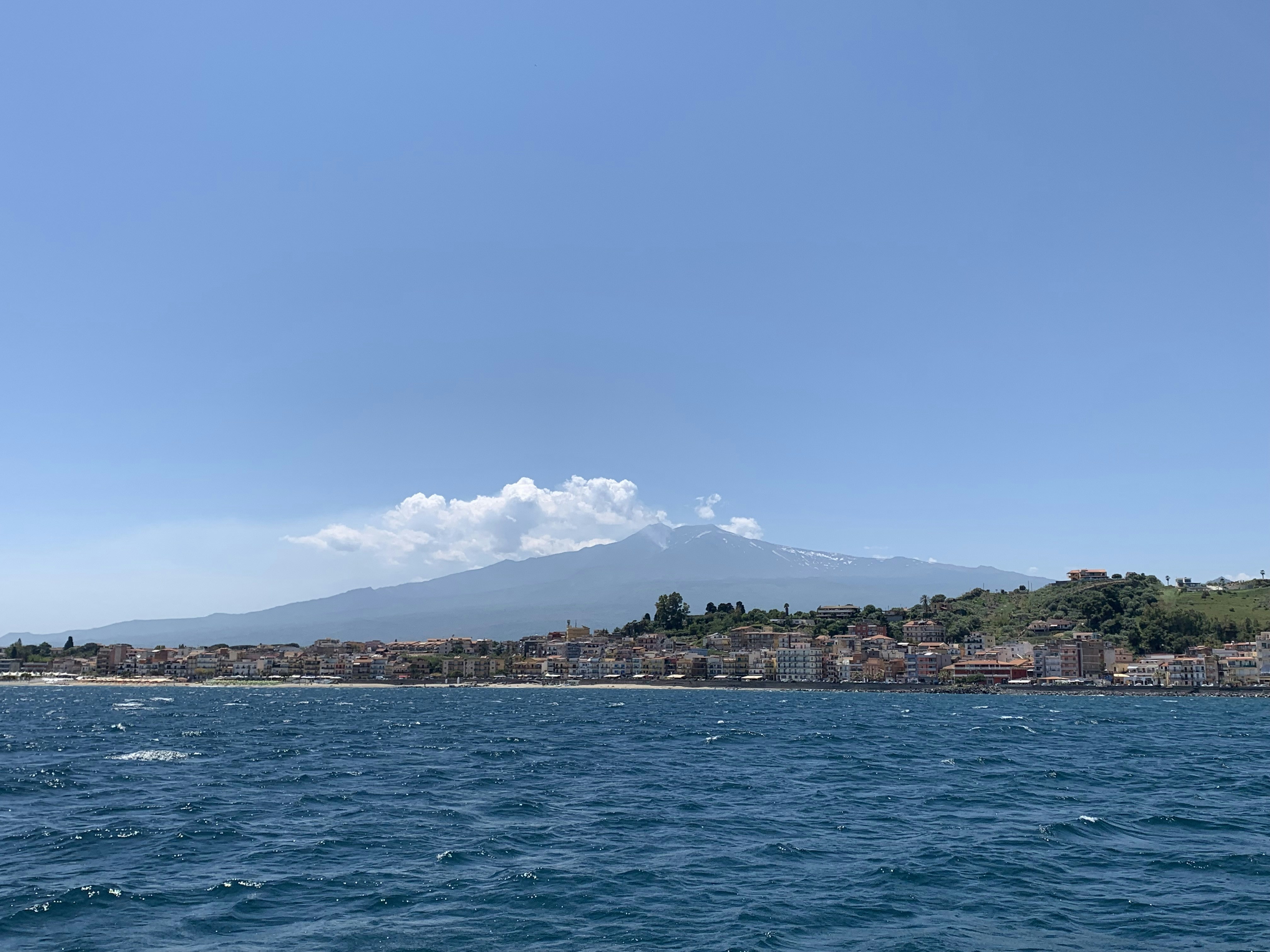 Snow-capped Mount Etna looms over a vibrant seaside town, with waves gently lapping at the shore under a clear blue sky.