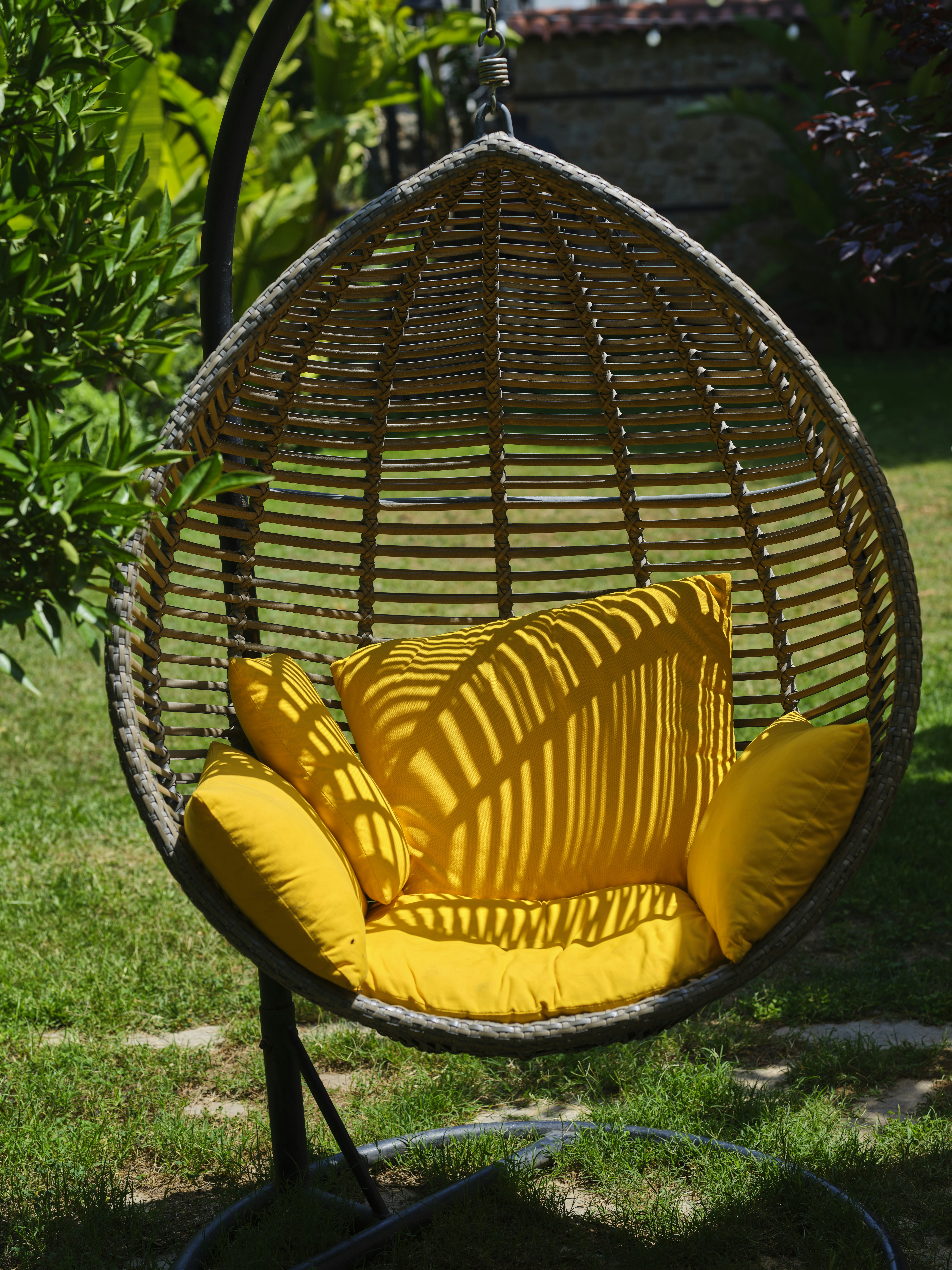 A suspended wicker chair adorned with vibrant yellow cushions, casting playful shadows on the grass in a tranquil garden setting.
