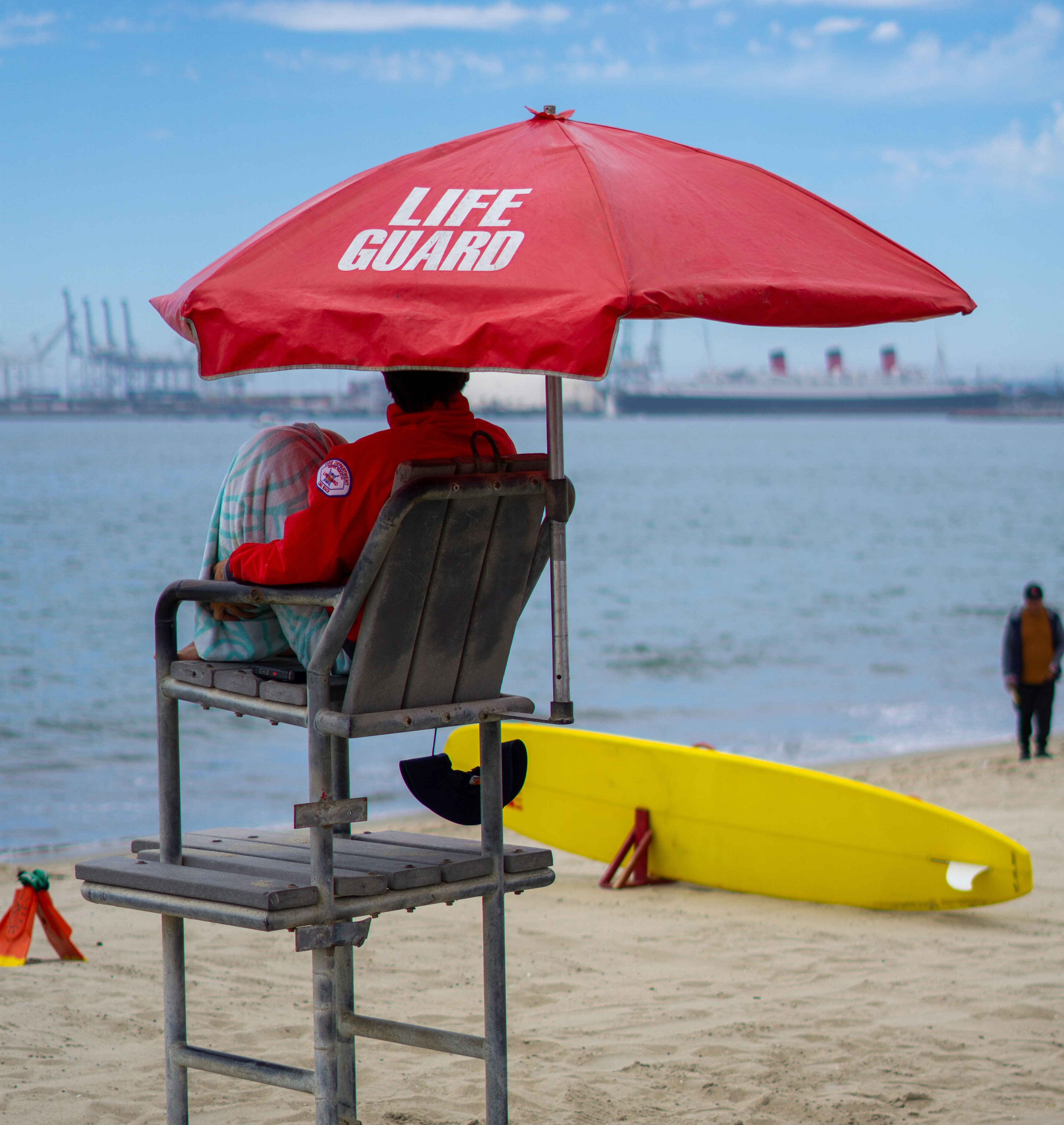 Lifeguard seated under a red umbrella, overlooking a sandy beach with a yellow surfboard nearby. The distant skyline features industrial structures and calm waters.