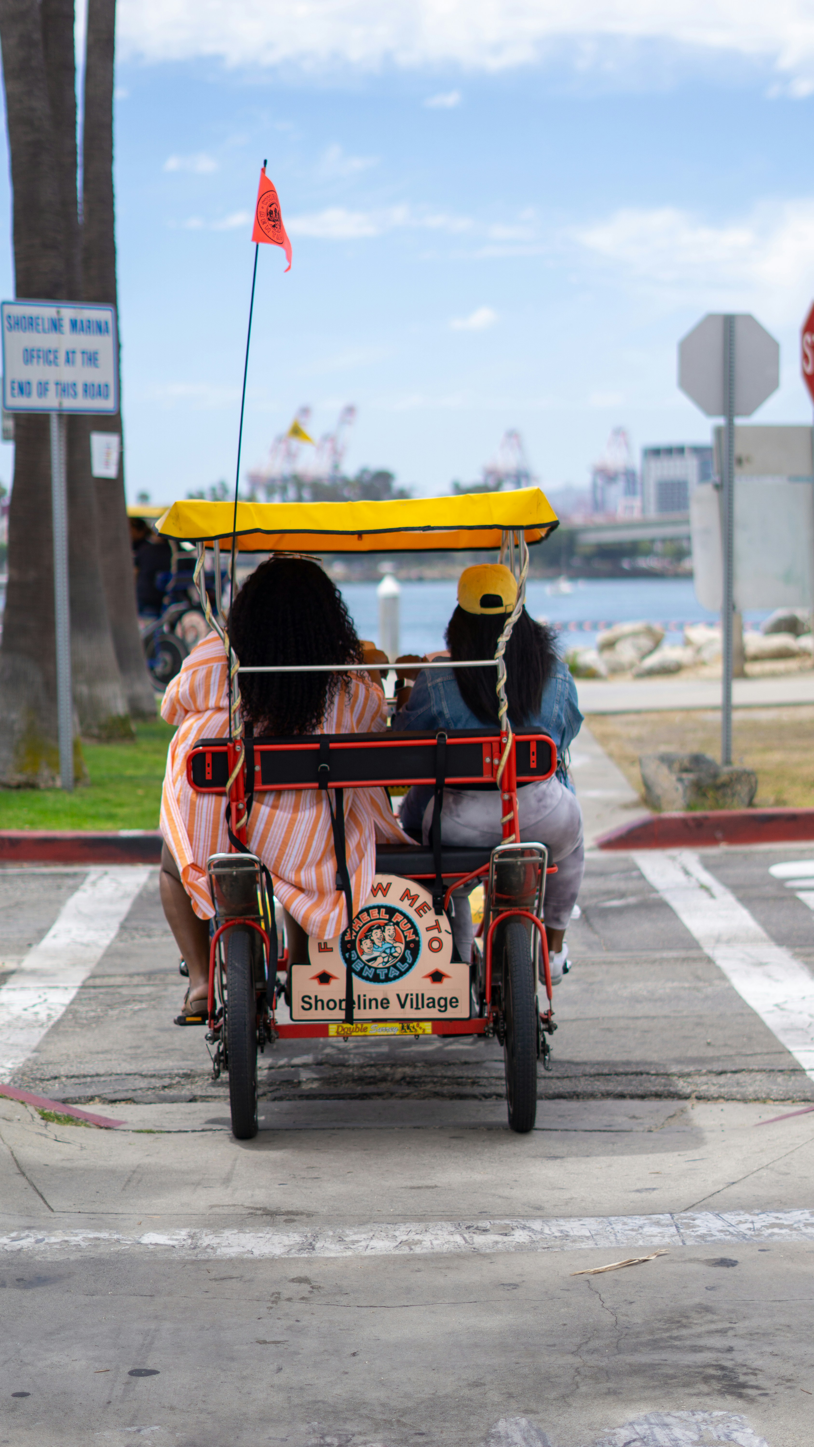Two friends enjoying a ride on a colorful pedal cart, with a view of the marina in the background. The scene captures a leisurely day out at Shoreline Village.