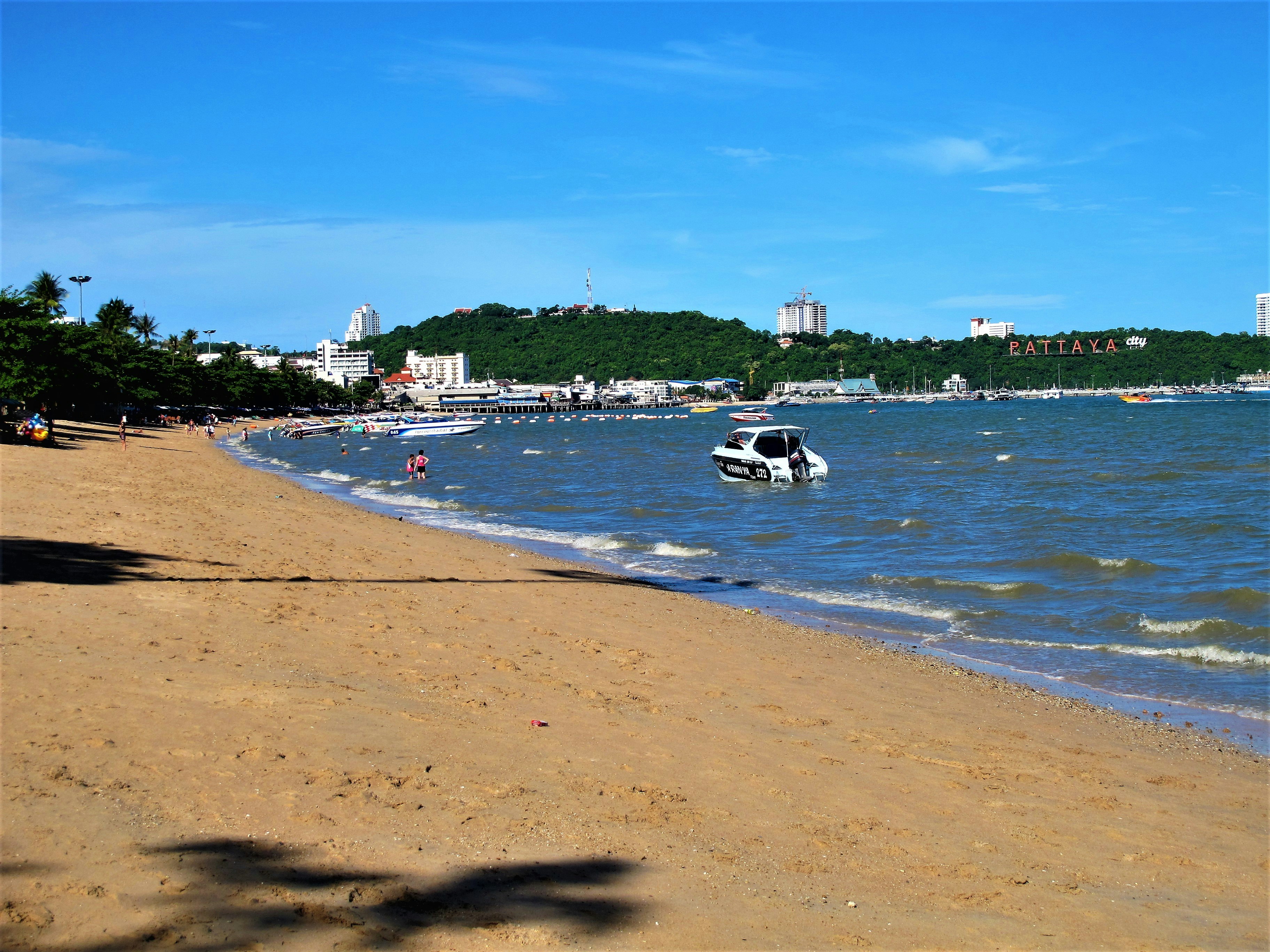 Sunlit beach with golden sand and gentle waves. A white motorboat cruises near the shore as distant buildings rise above a green hill.
