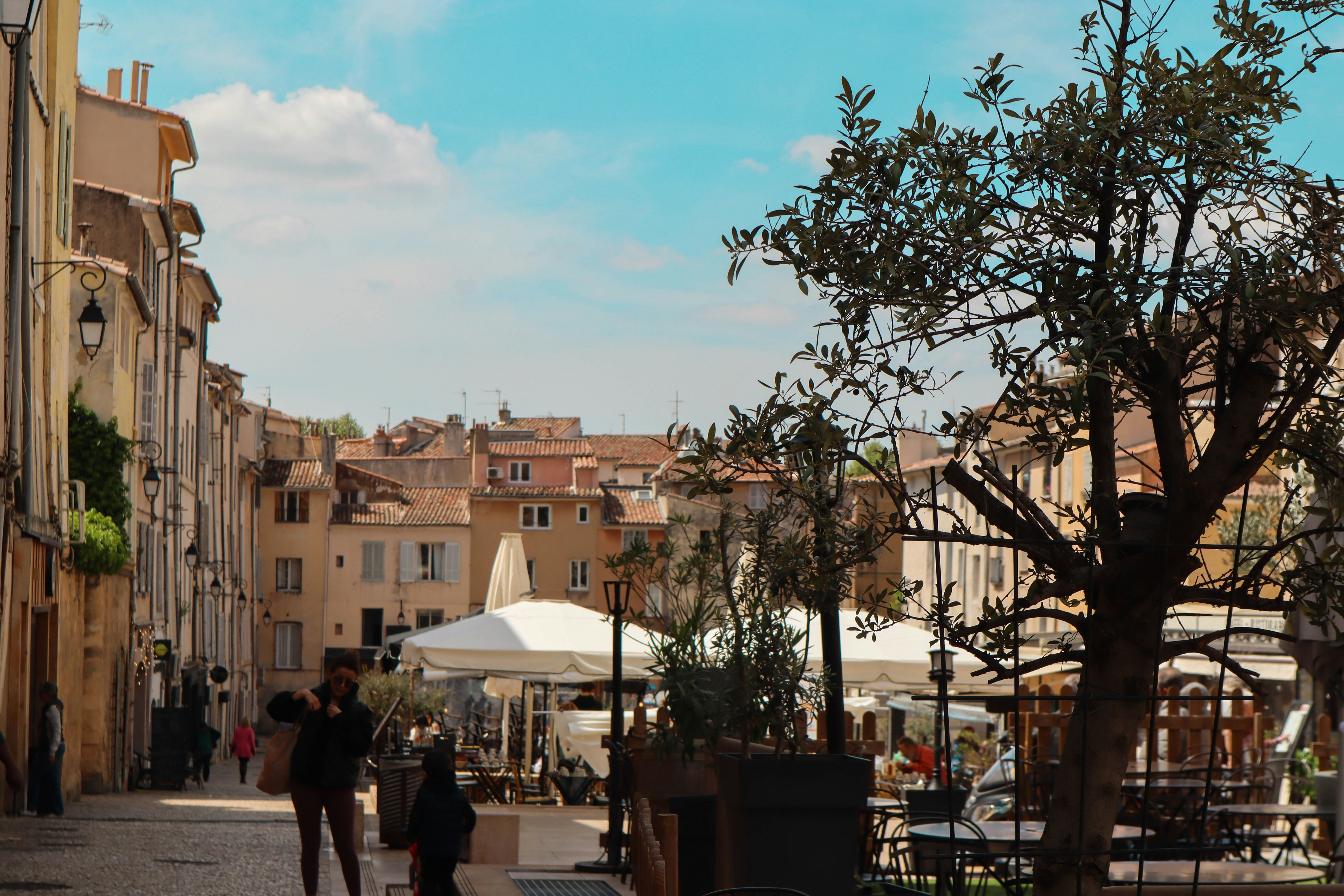 People walking through a sunlit European square with outdoor cafes and historic buildings under a clear blue sky.